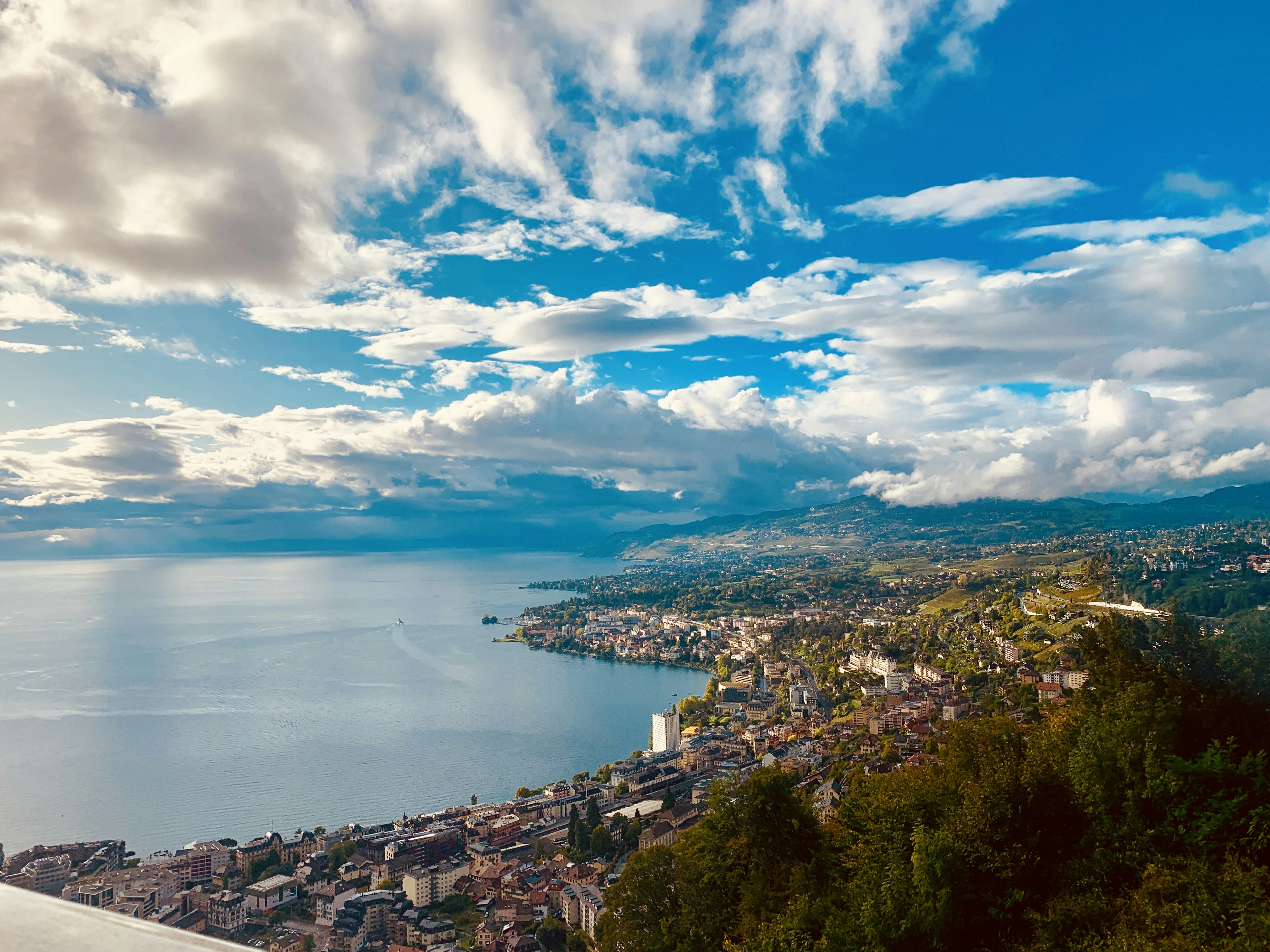 A panoramic view of Lake Geneva and its surrounding landscape, showcasing a blend of urban and natural beauty. The clouds reflect a soft light over the water's surface.