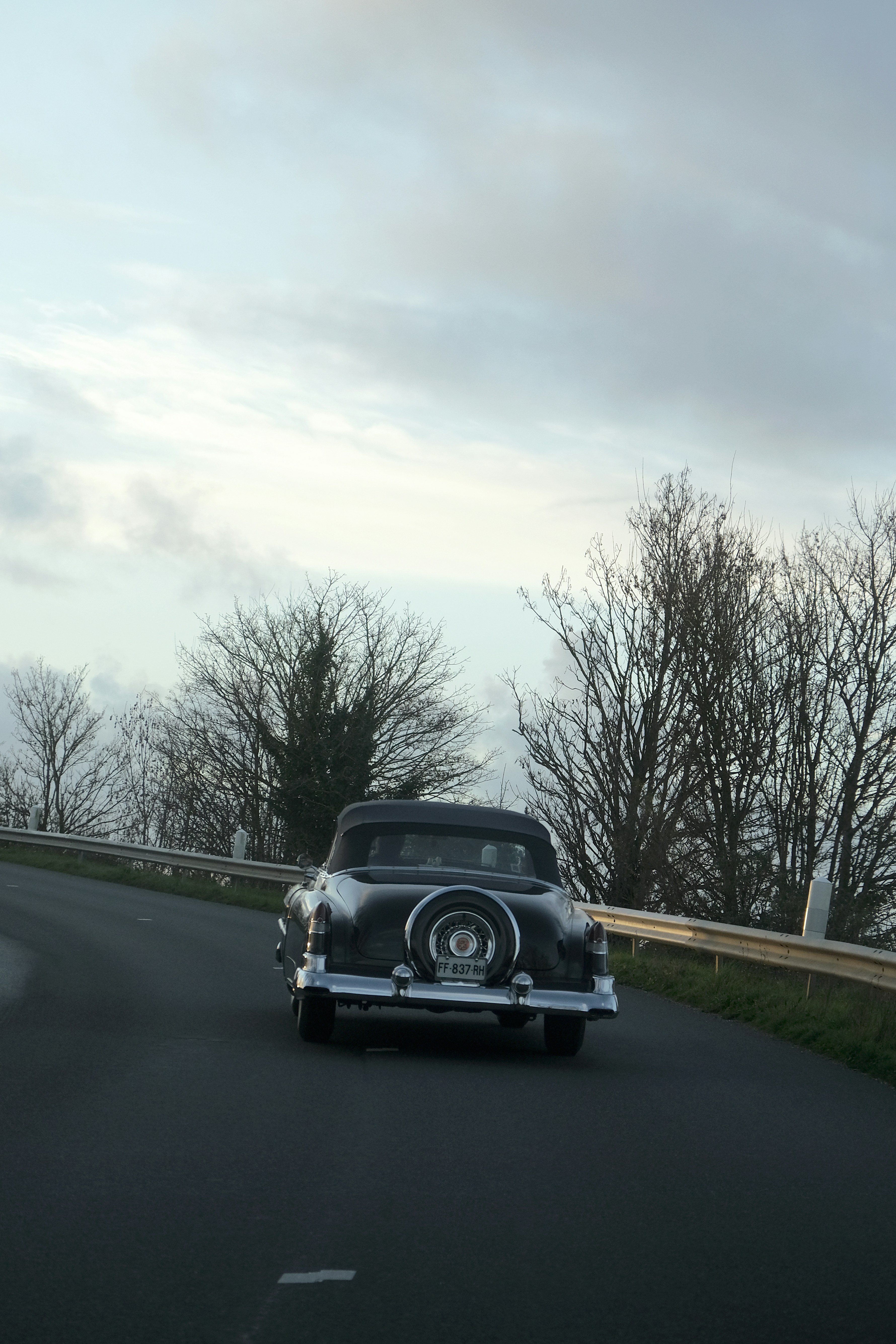 Classic convertible car navigating a winding road under a cloudy sky, surrounded by leafless trees.
