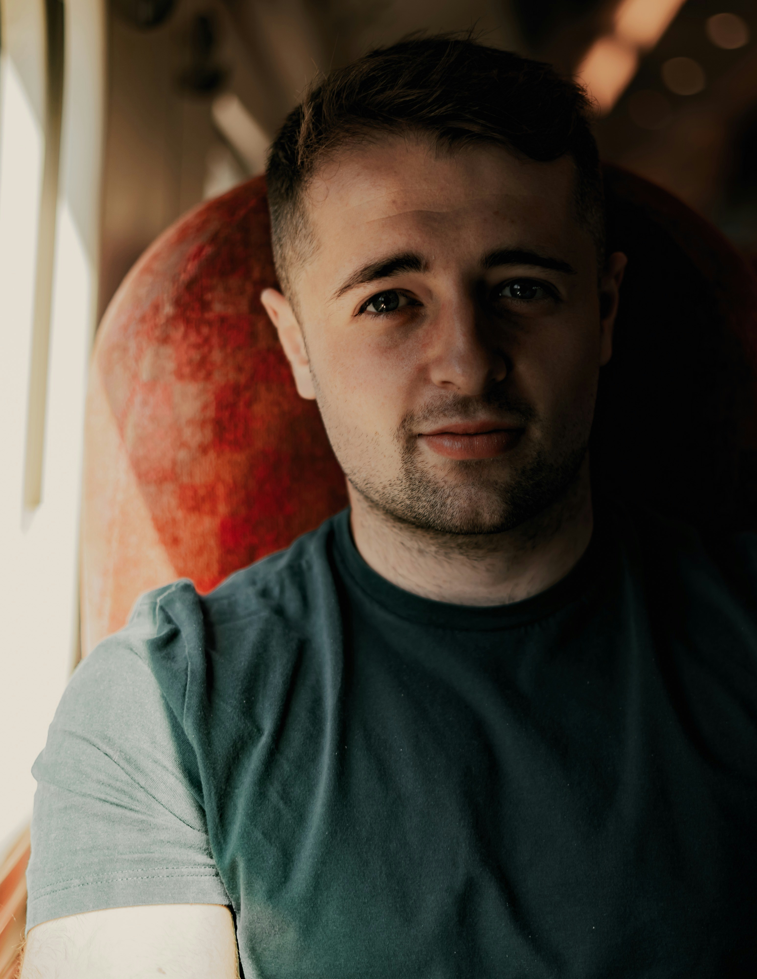 Young man seated by a window on a train, illuminated by soft natural light, with a textured red seat in the background.
