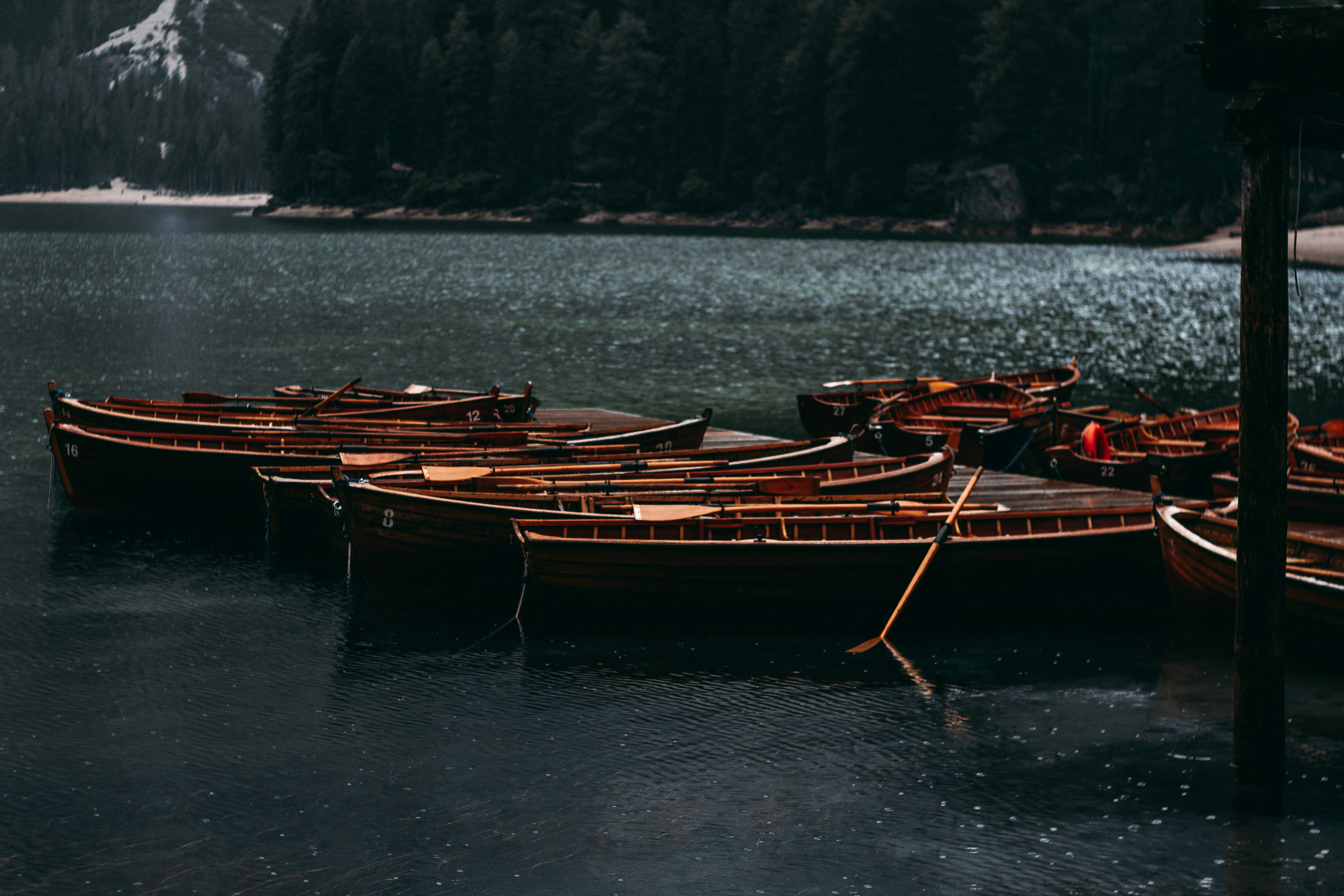 A collection of wooden boats gently resting on a tranquil lake, surrounded by dense forest and misty mountains. The scene conveys a peaceful atmosphere.