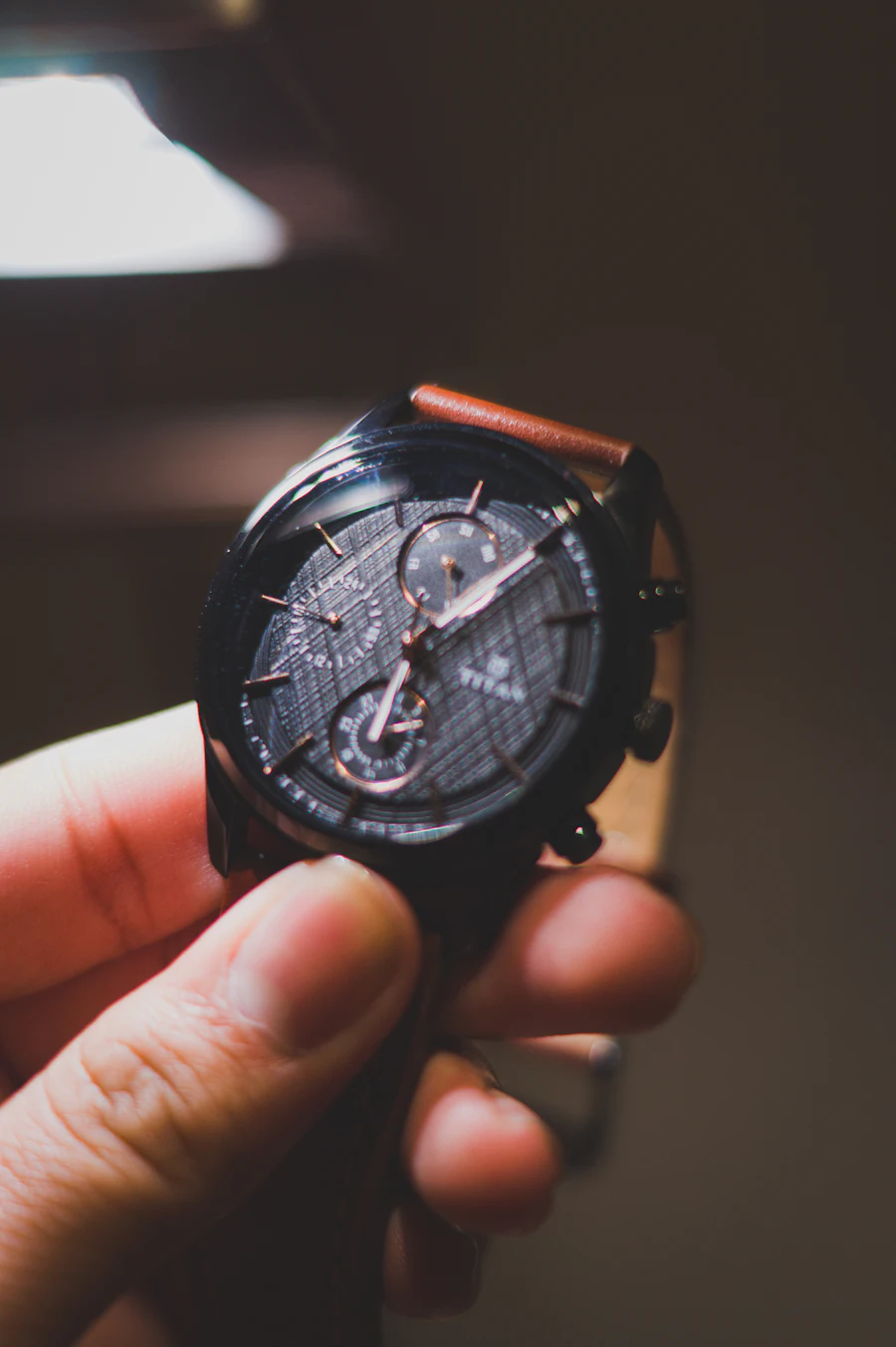 A hand holds a black wristwatch with a textured dial and brown strap in soft window light. The background falls into a warm blur.