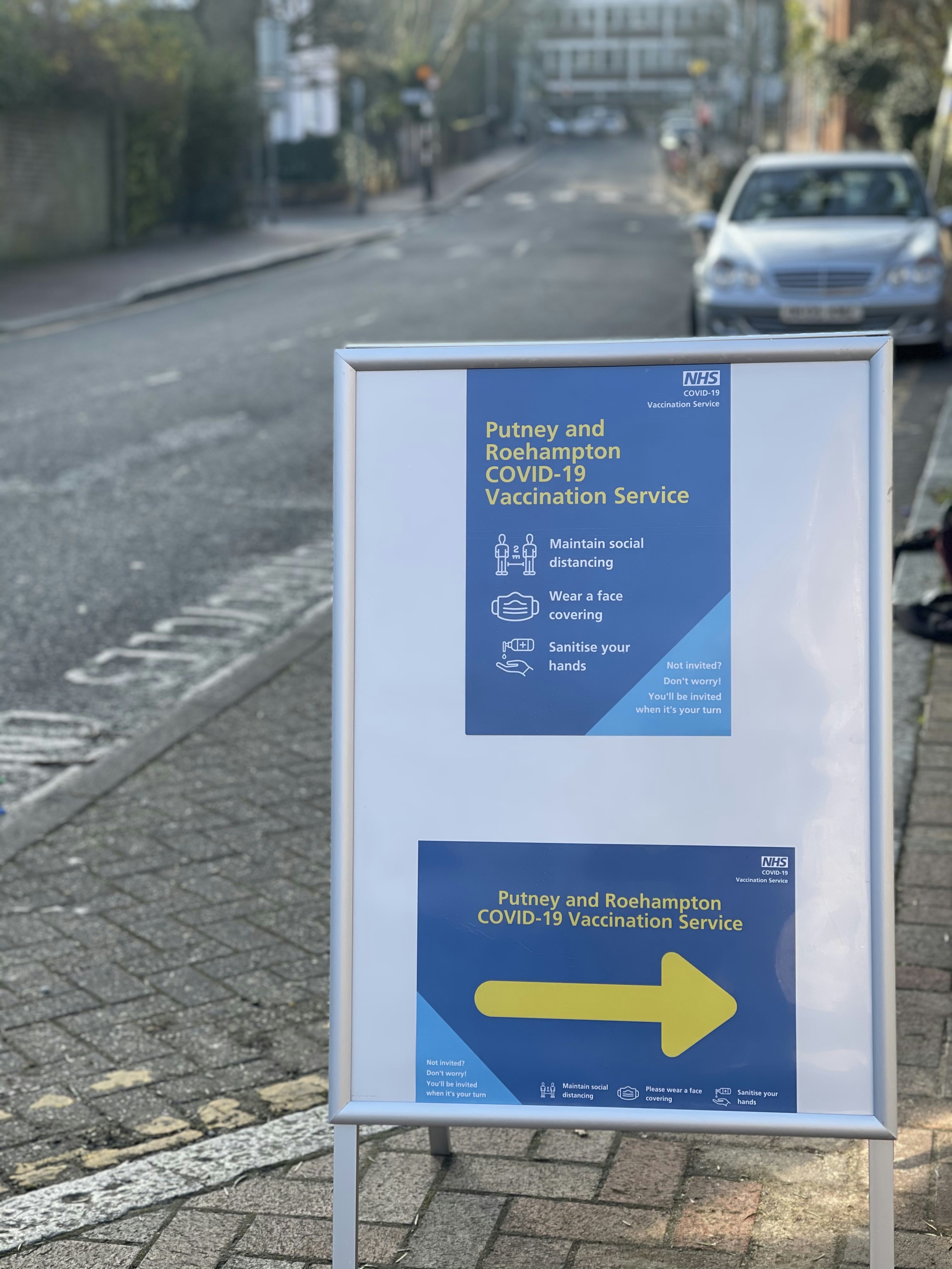 Double-panel public information sign on a sidewalk displays blue COVID-19 vaccination posters. The street behind is quiet, with a car parked and a blurred cityscape.