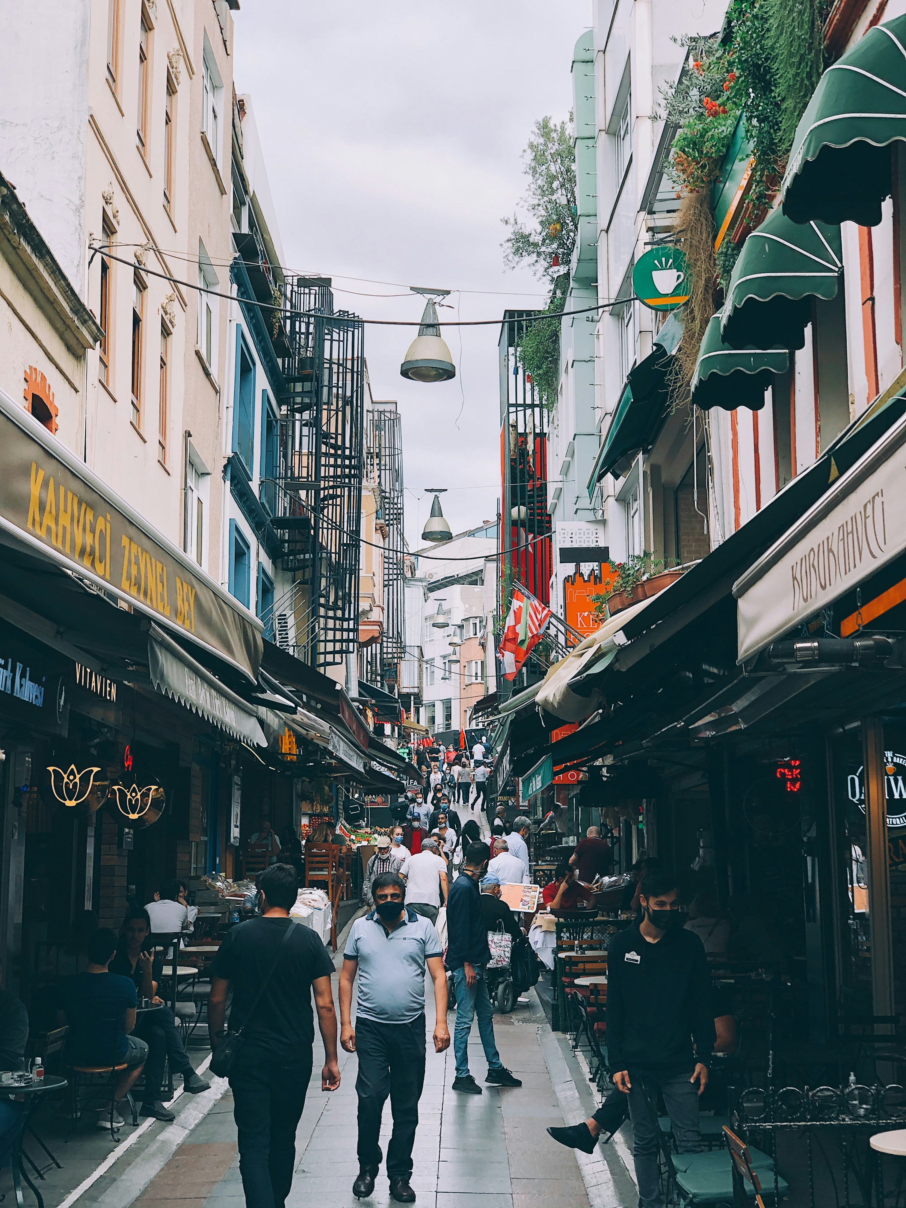 Bustling street scene in a vibrant urban area, showcasing cafes and shops lined with patrons and greenery above.