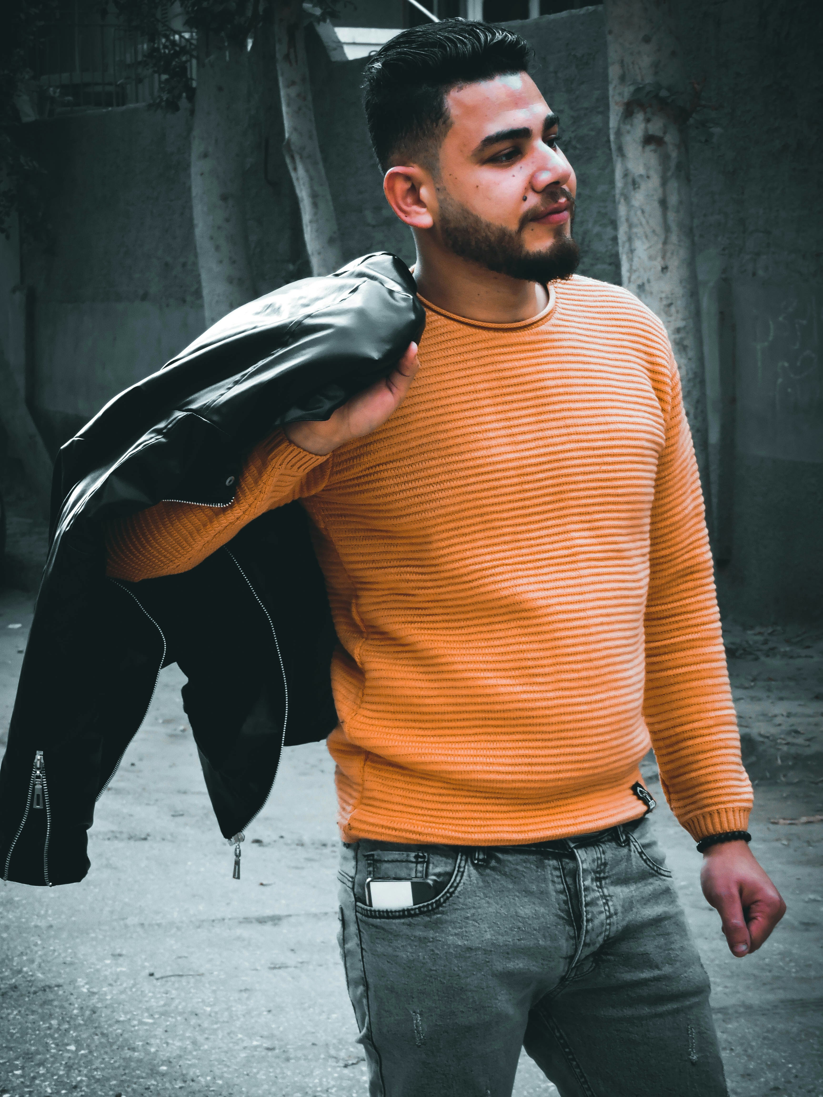 Young man in an orange striped sweater confidently holds a leather jacket over his shoulder while standing on a city street.