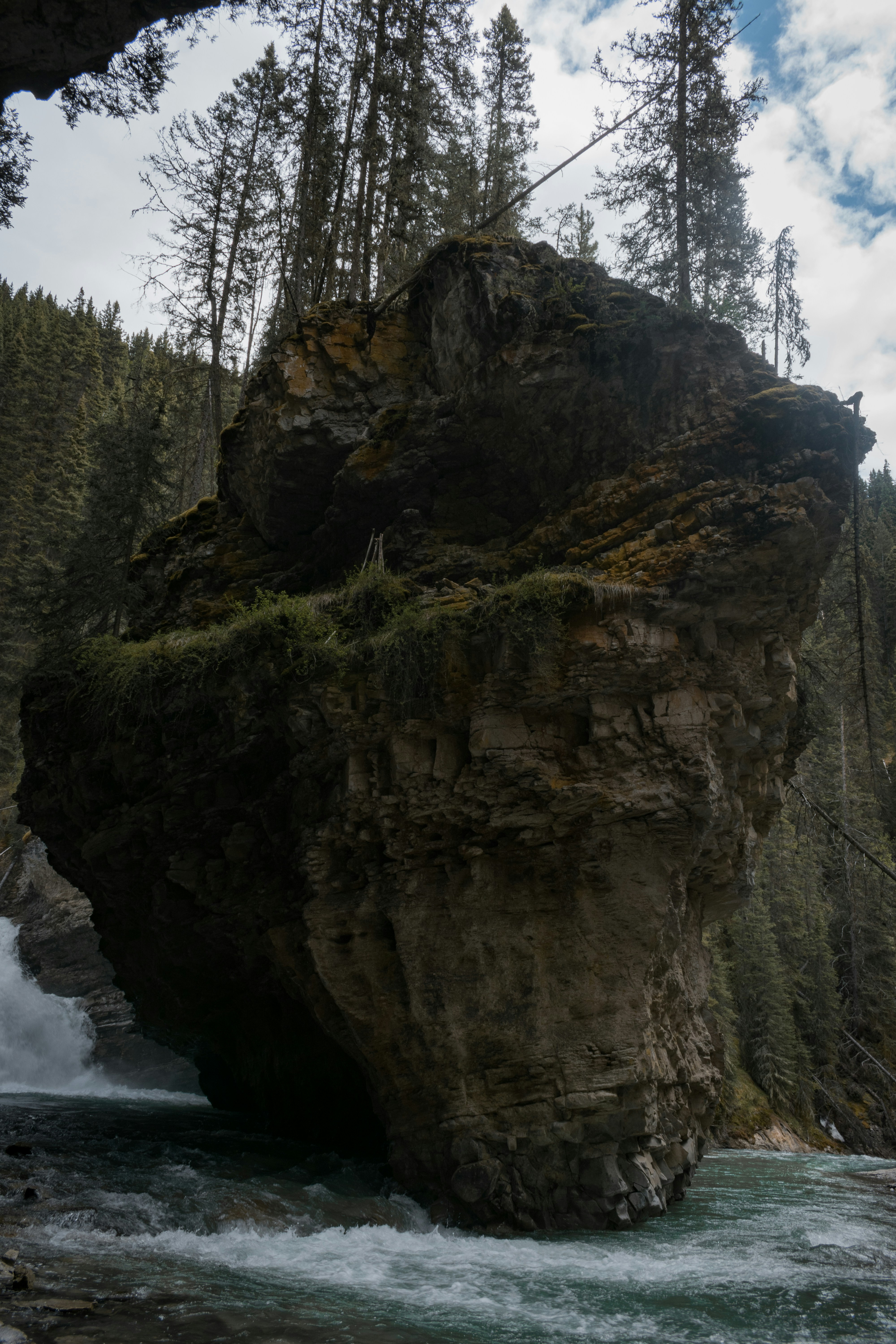 A large rock formation stands majestically beside a rushing river, surrounded by tall trees under a cloudy sky.
