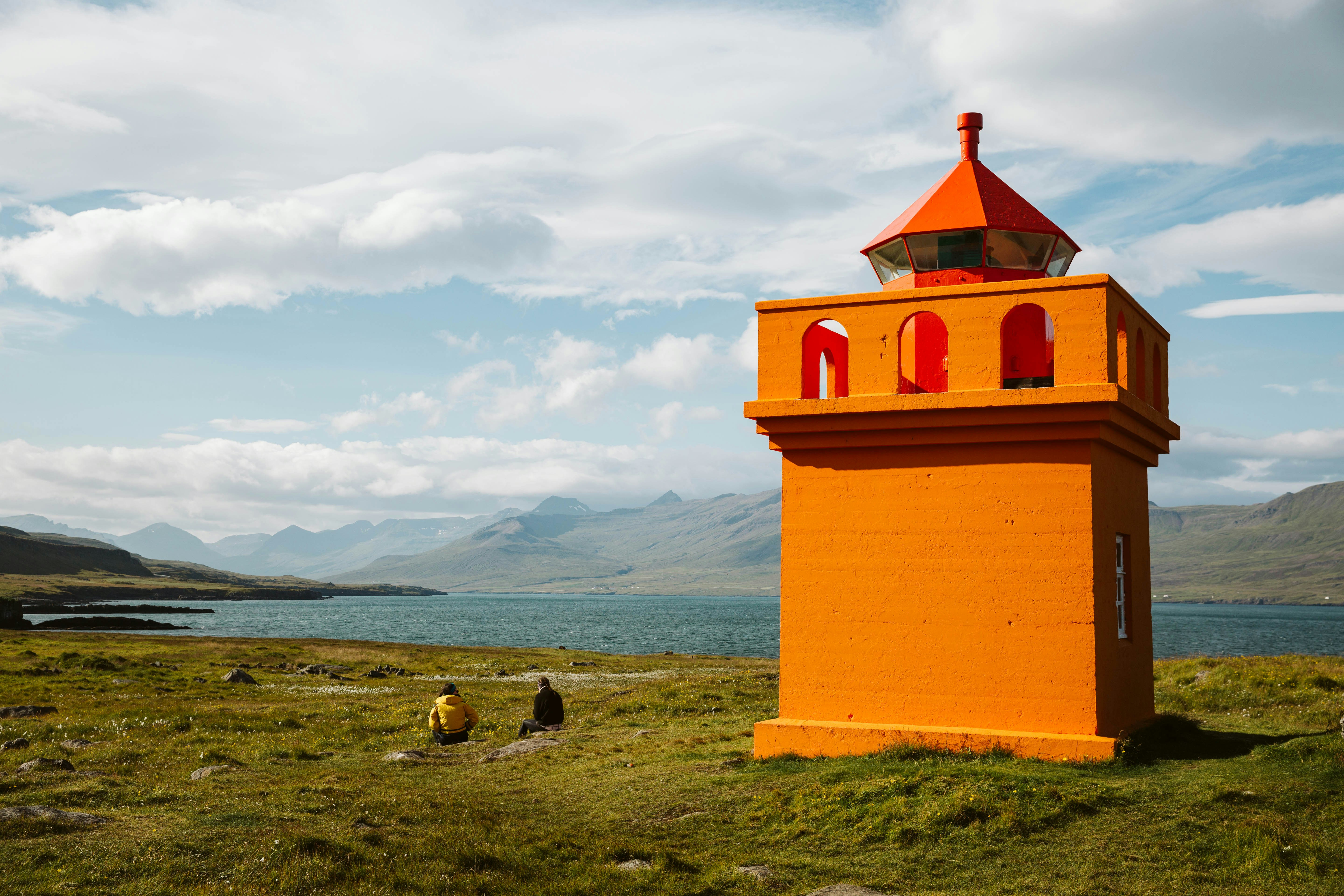 Bright orange lighthouse stands against a serene landscape, with two figures seated nearby, gazing at the tranquil waters and distant mountains.