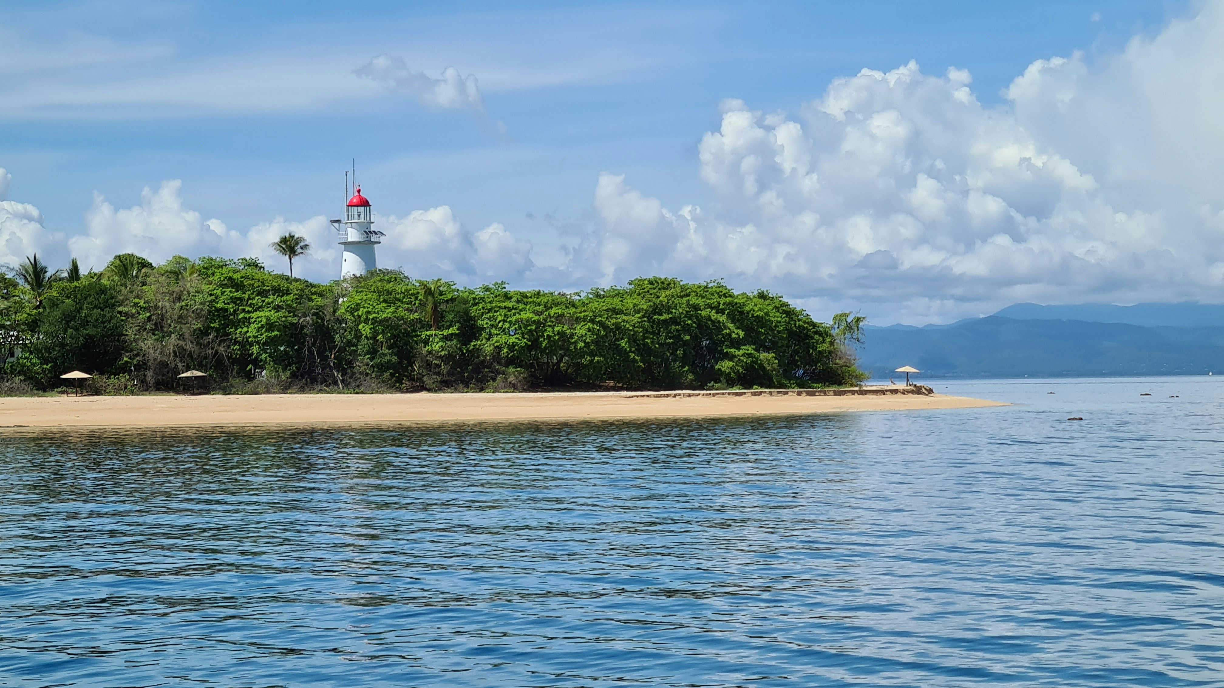 Lighthouse with red top stands among lush greenery on a sandy island under a bright blue sky.