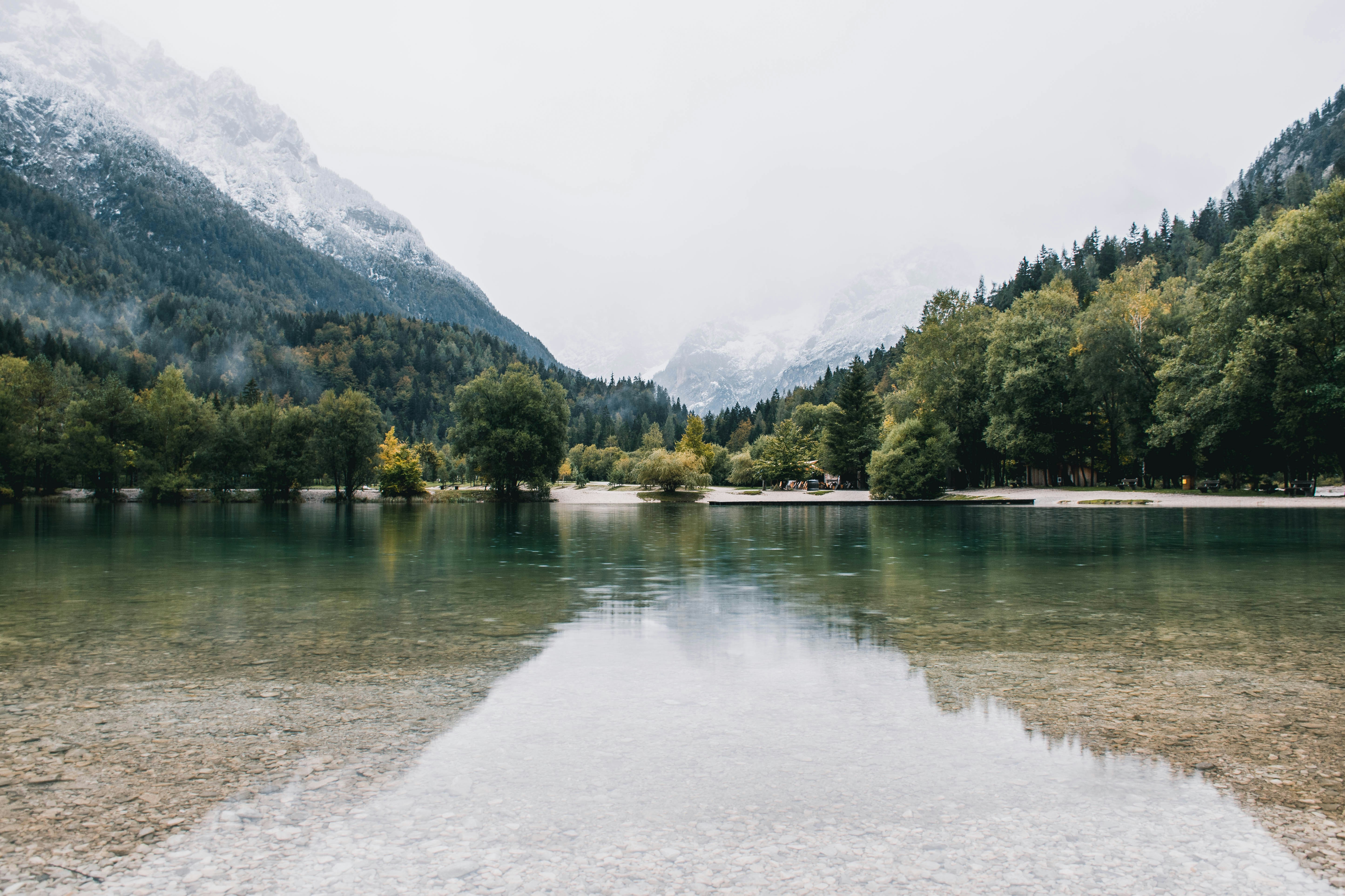 Clear alpine lake mirroring lush trees and snow-capped mountains under a cloudy sky.