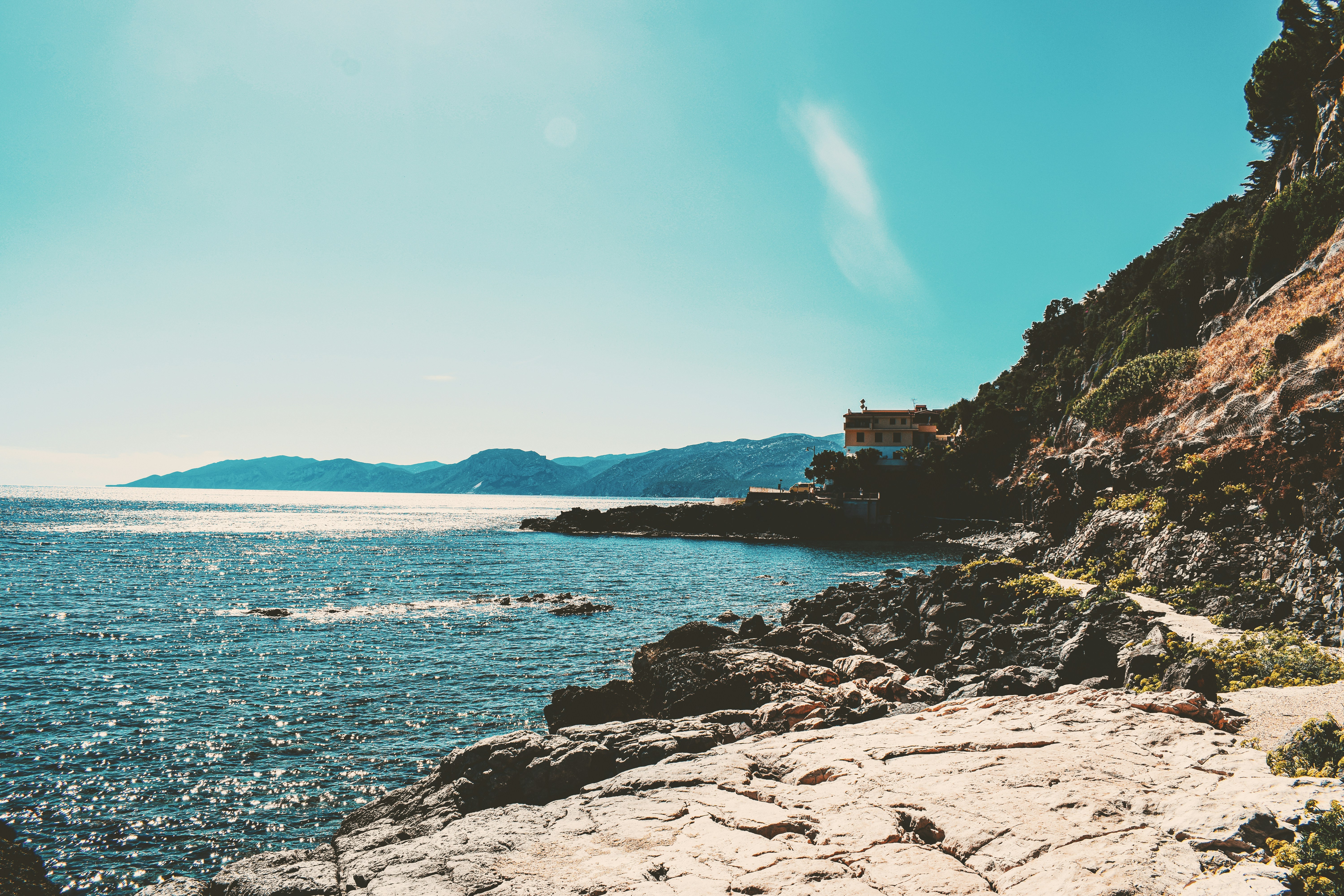 Rocky coastline with clear blue waters and distant hills under a bright sky.
