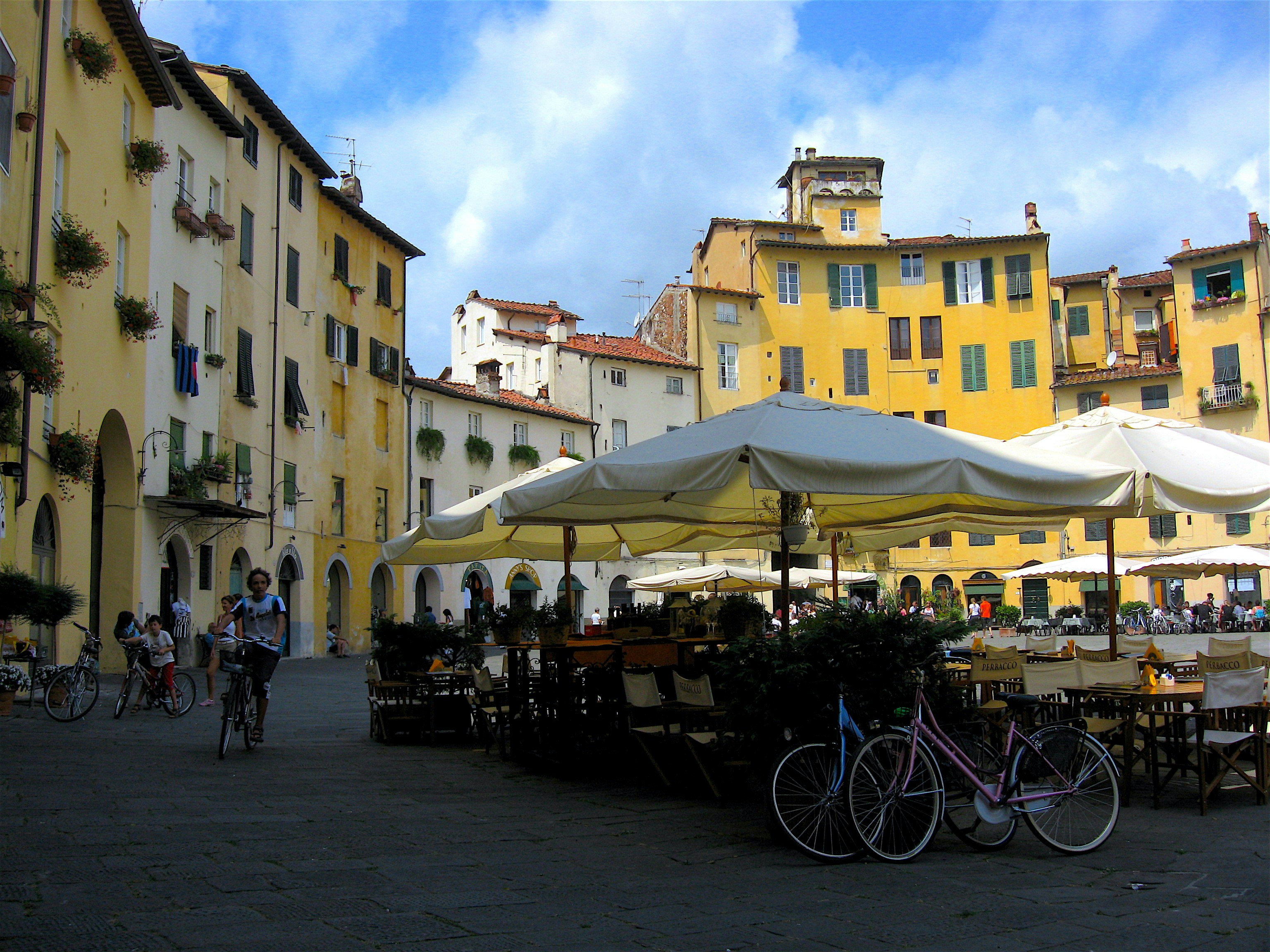 Colorful buildings surround a lively Italian square with bicycles parked near outdoor cafes under large umbrellas.