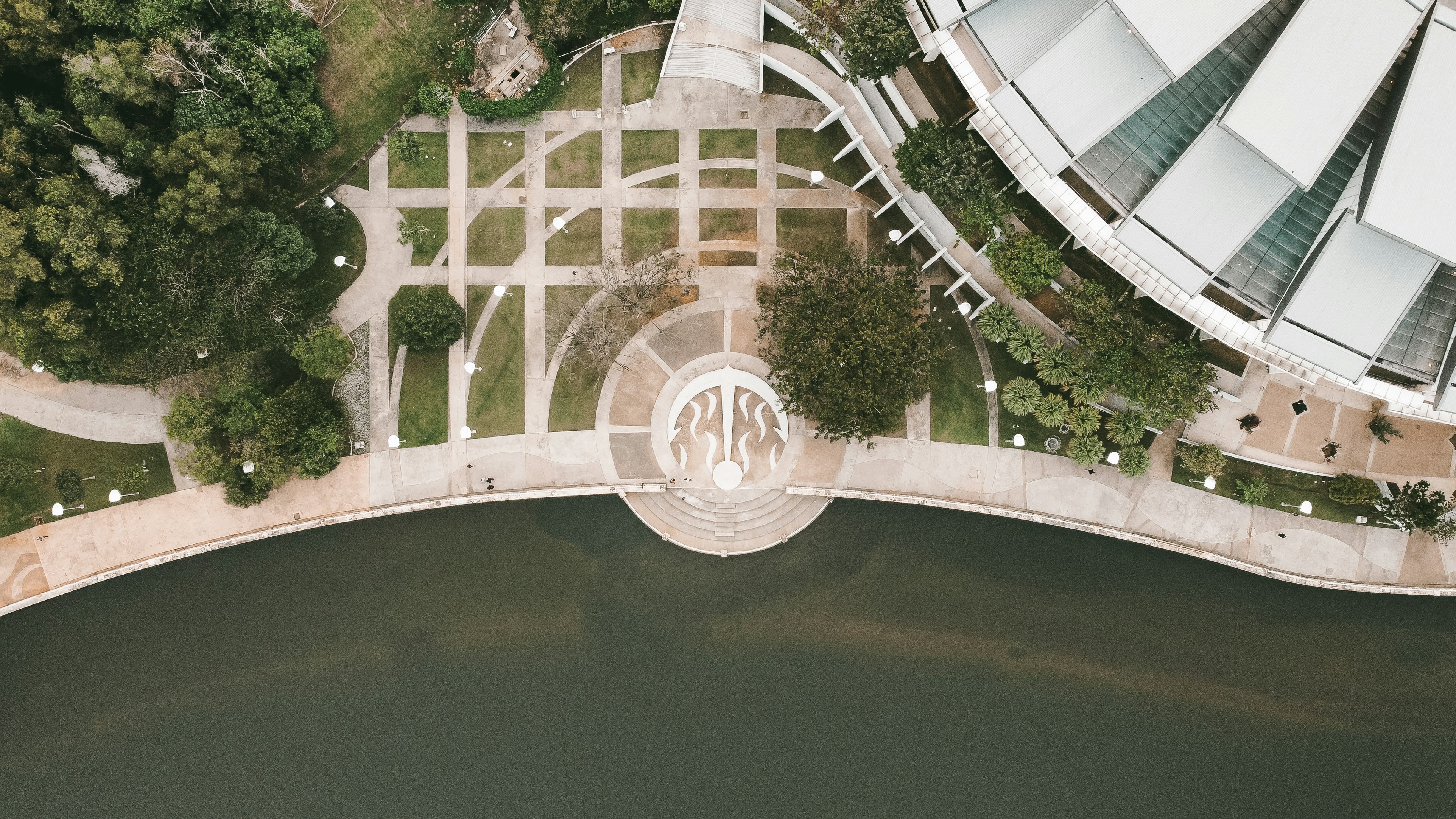 Aerial perspective of a park with geometric pathways and a central circular design by the water's edge, surrounded by lush greenery and modern architecture.