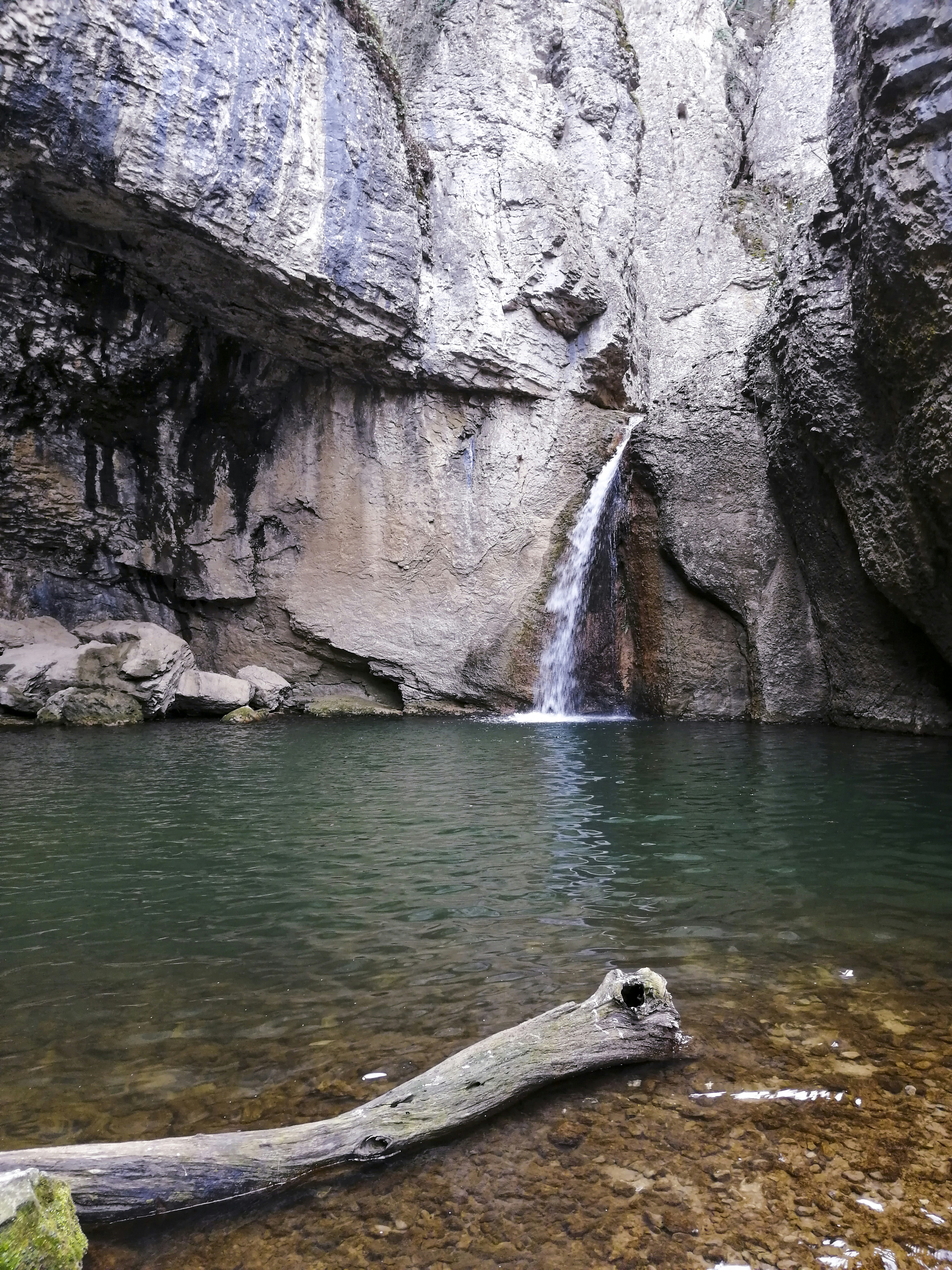 A serene waterfall cascades into a tranquil pool surrounded by rugged cliffs and a fallen log in the foreground.