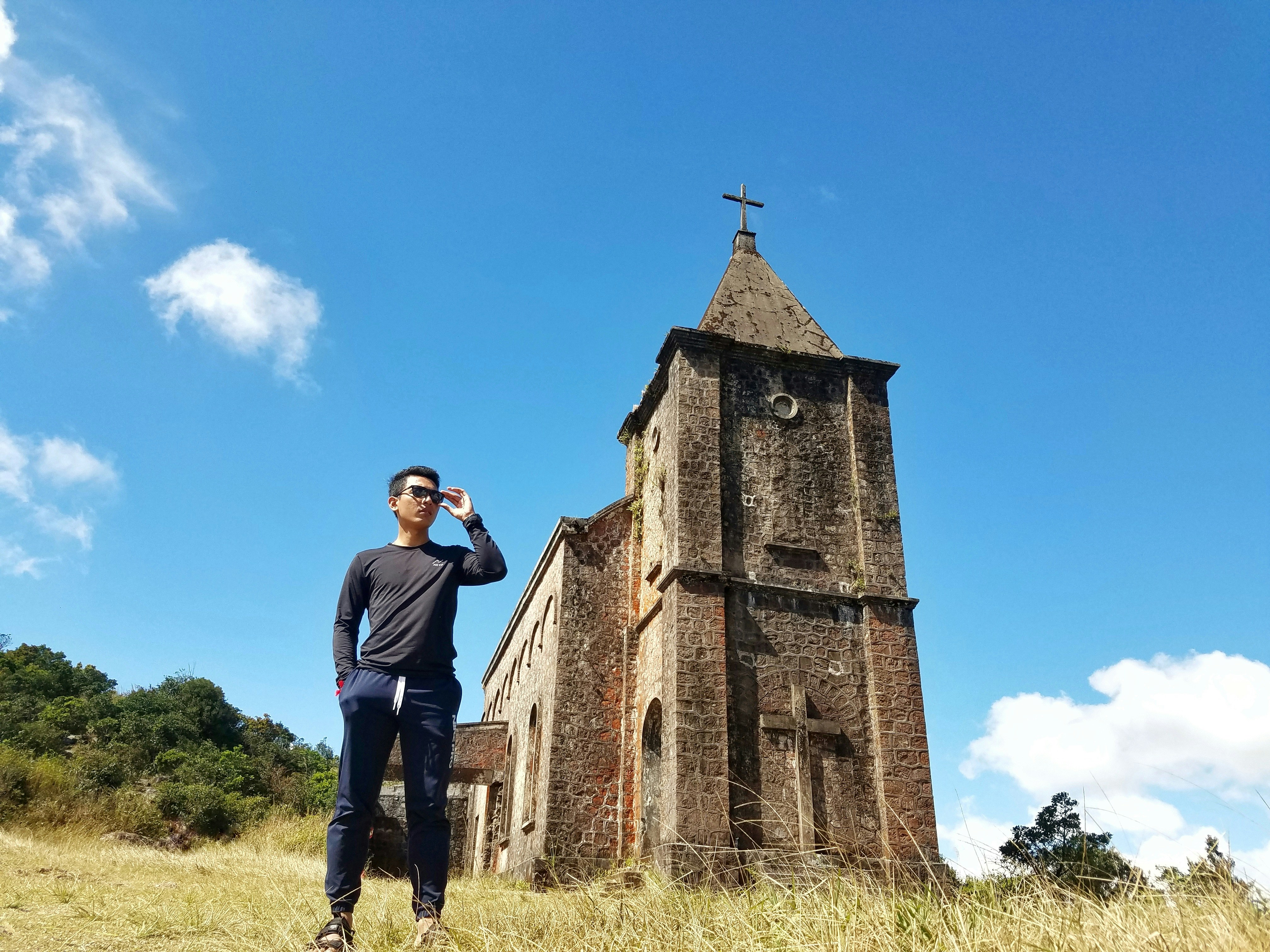 A man stands beside an old stone church under a bright blue sky; the scene emphasizes a lone visitor against the rugged architecture and open landscape.