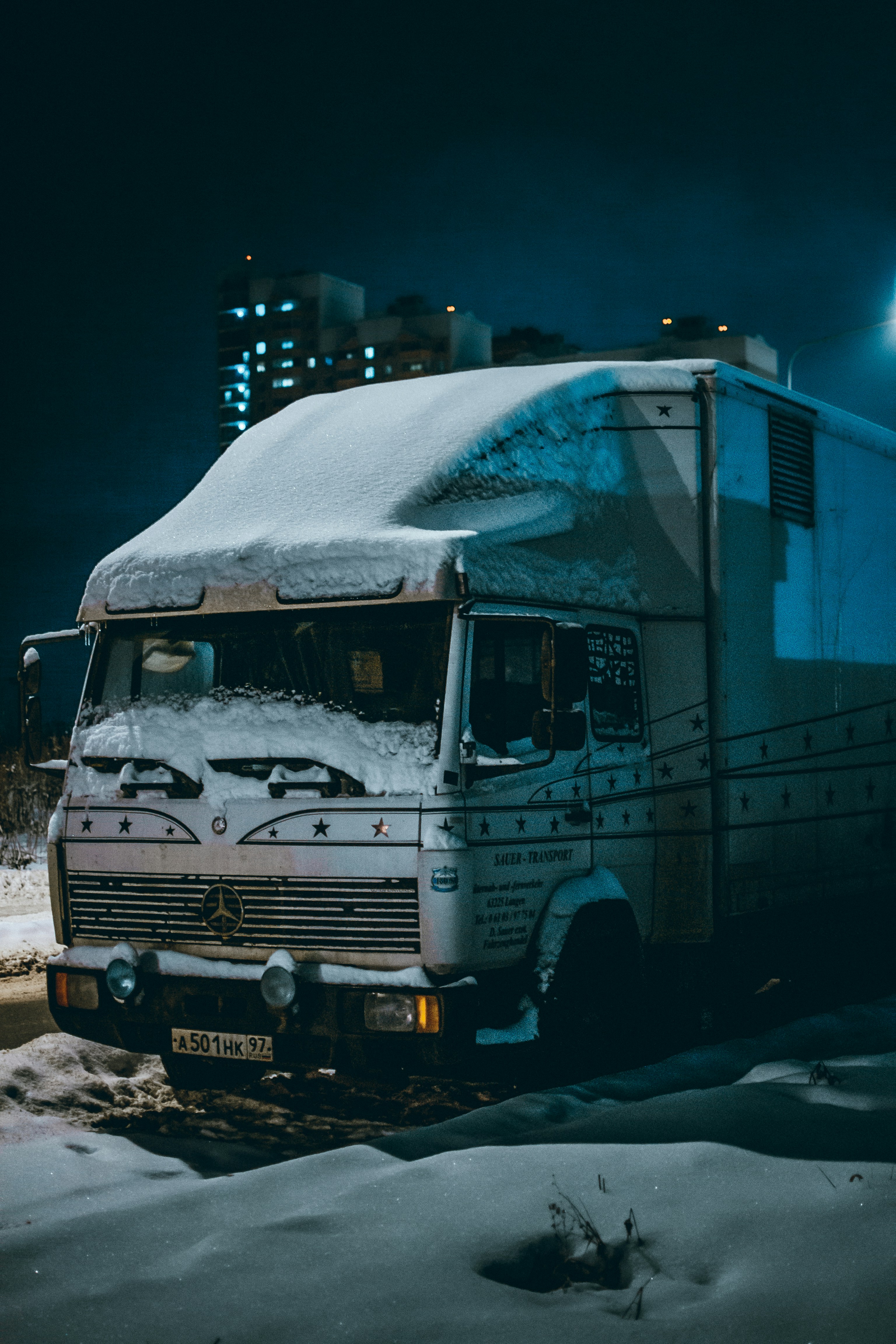A vintage truck covered in snow, parked on a quiet street, illuminated by city lights in the background.