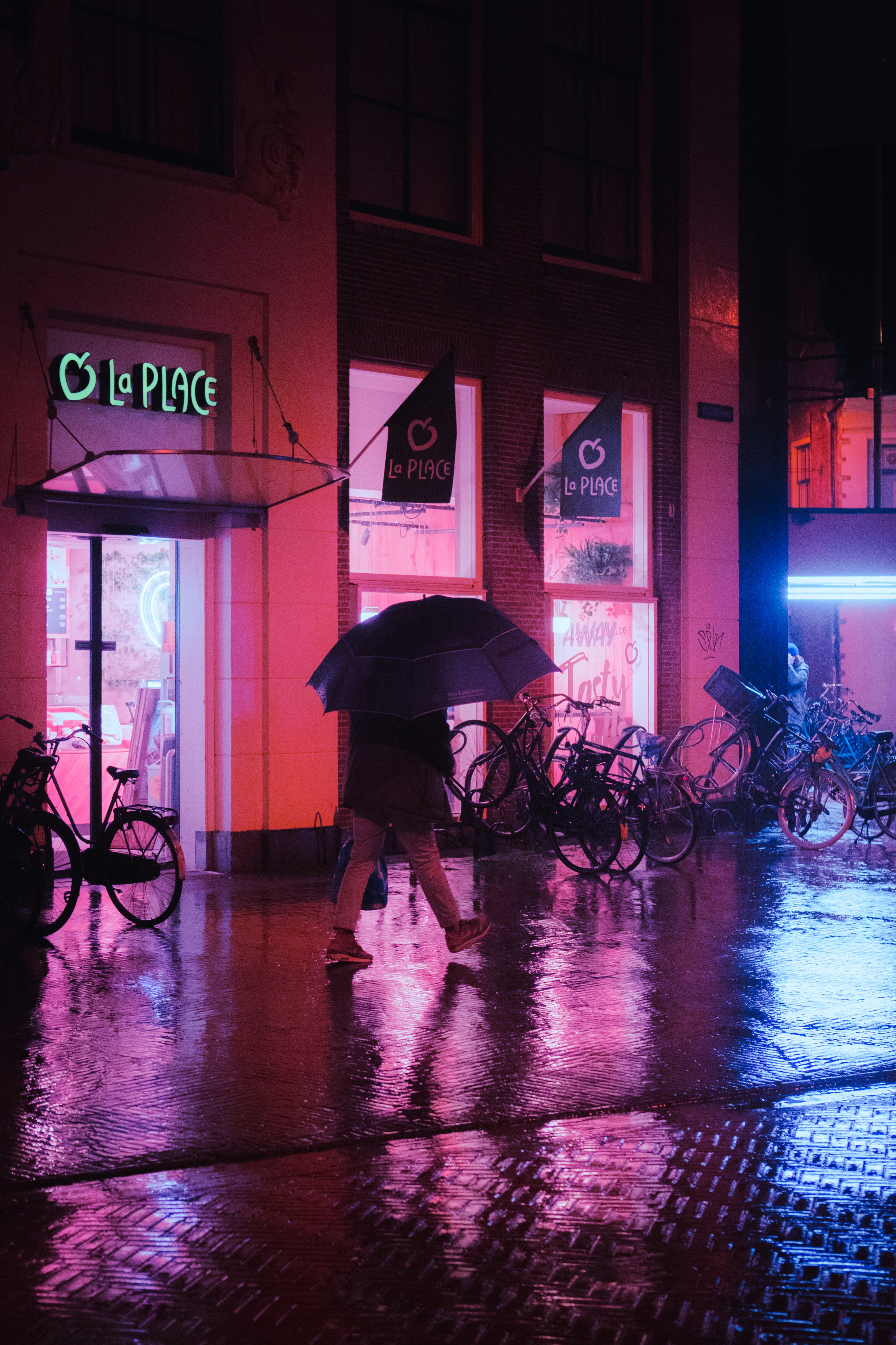 A solitary figure with an umbrella walks along a rain-soaked street, illuminated by vibrant neon lights from nearby shops. The wet pavement reflects the colorful glow, enhancing the urban atmosphere.