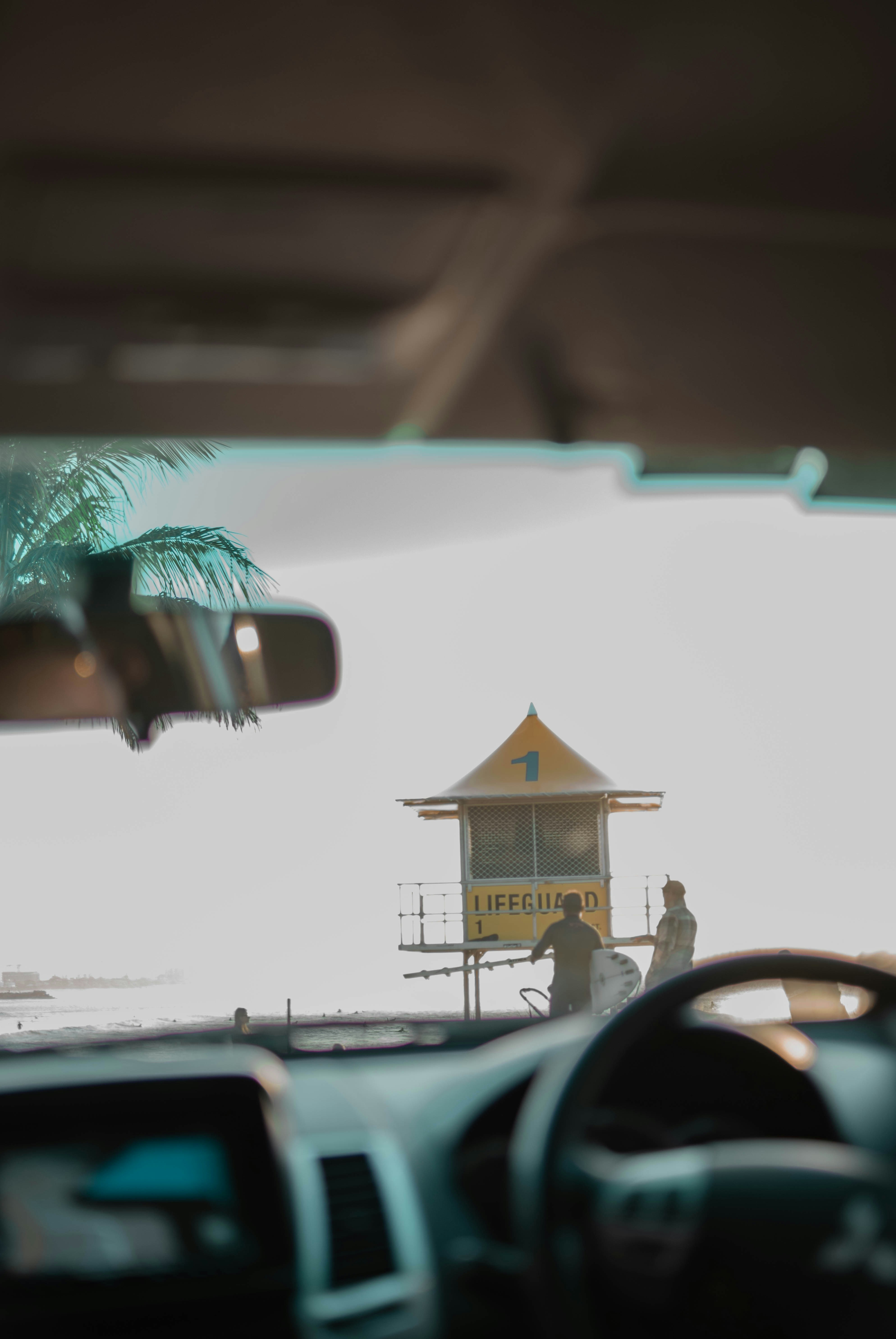 Lifeguard station stands prominently by the beach, framed by the interior of a vehicle. Two figures approach the station, hinting at a day of vigilance by the water.
