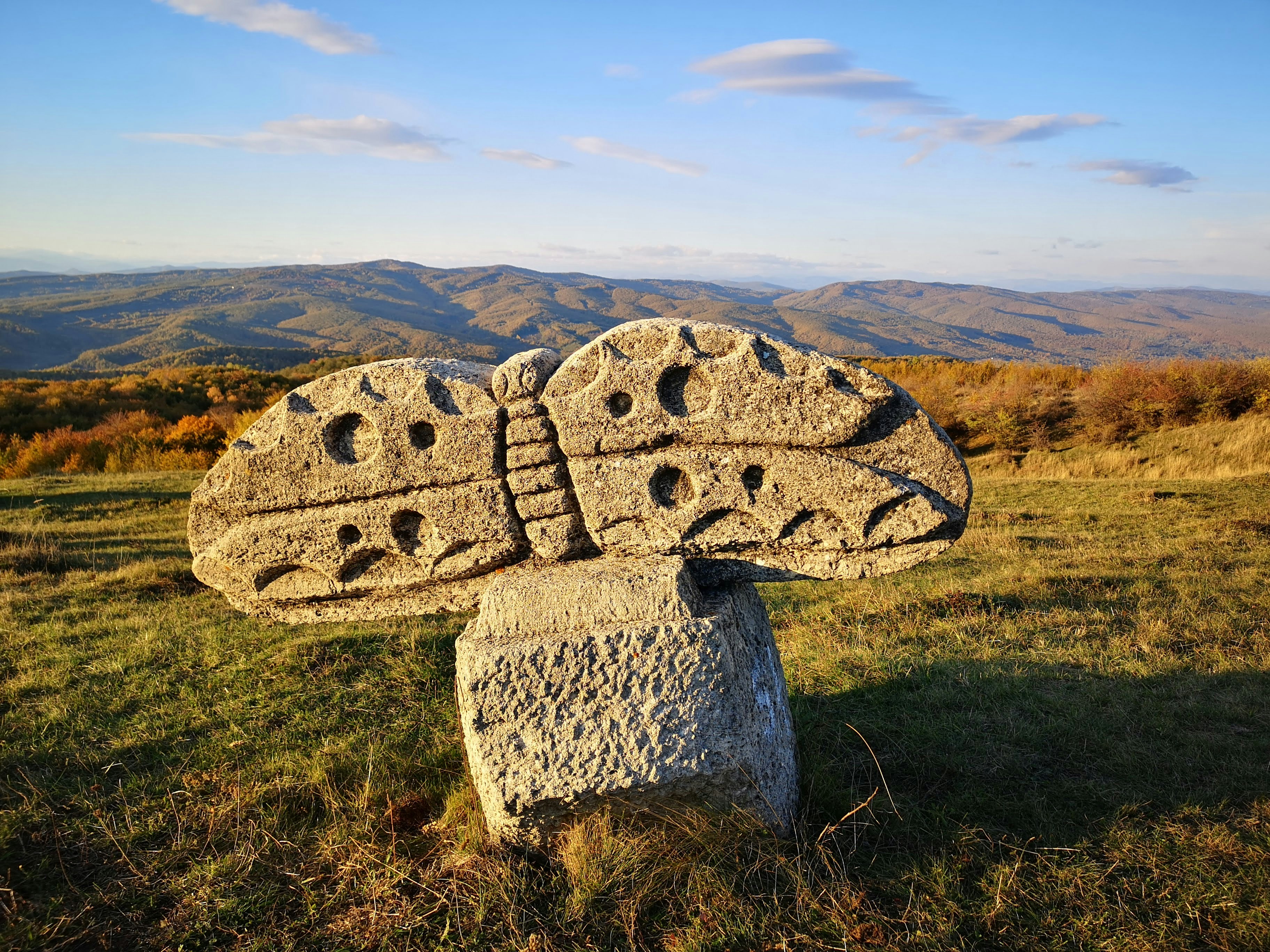 Granite sculpture of a butterfly perched on a stone base, set against a backdrop of rolling hills and autumn foliage. The warm light enhances the natural beauty of the landscape.