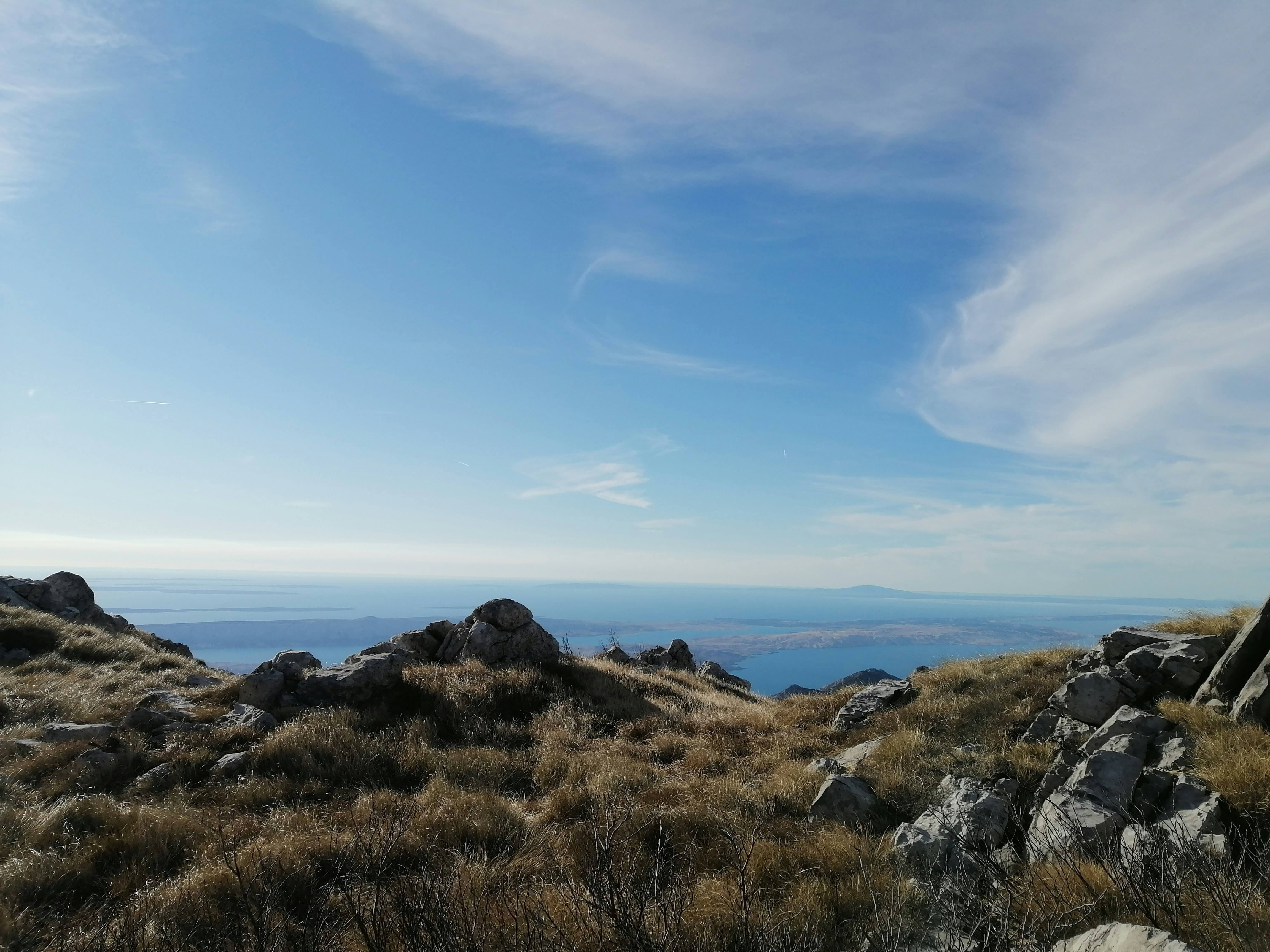 Rocky coastal ridge with dry grasses leads to a calm sea at the horizon under a clear blue sky. The scene emphasizes open landscape and natural textures.
