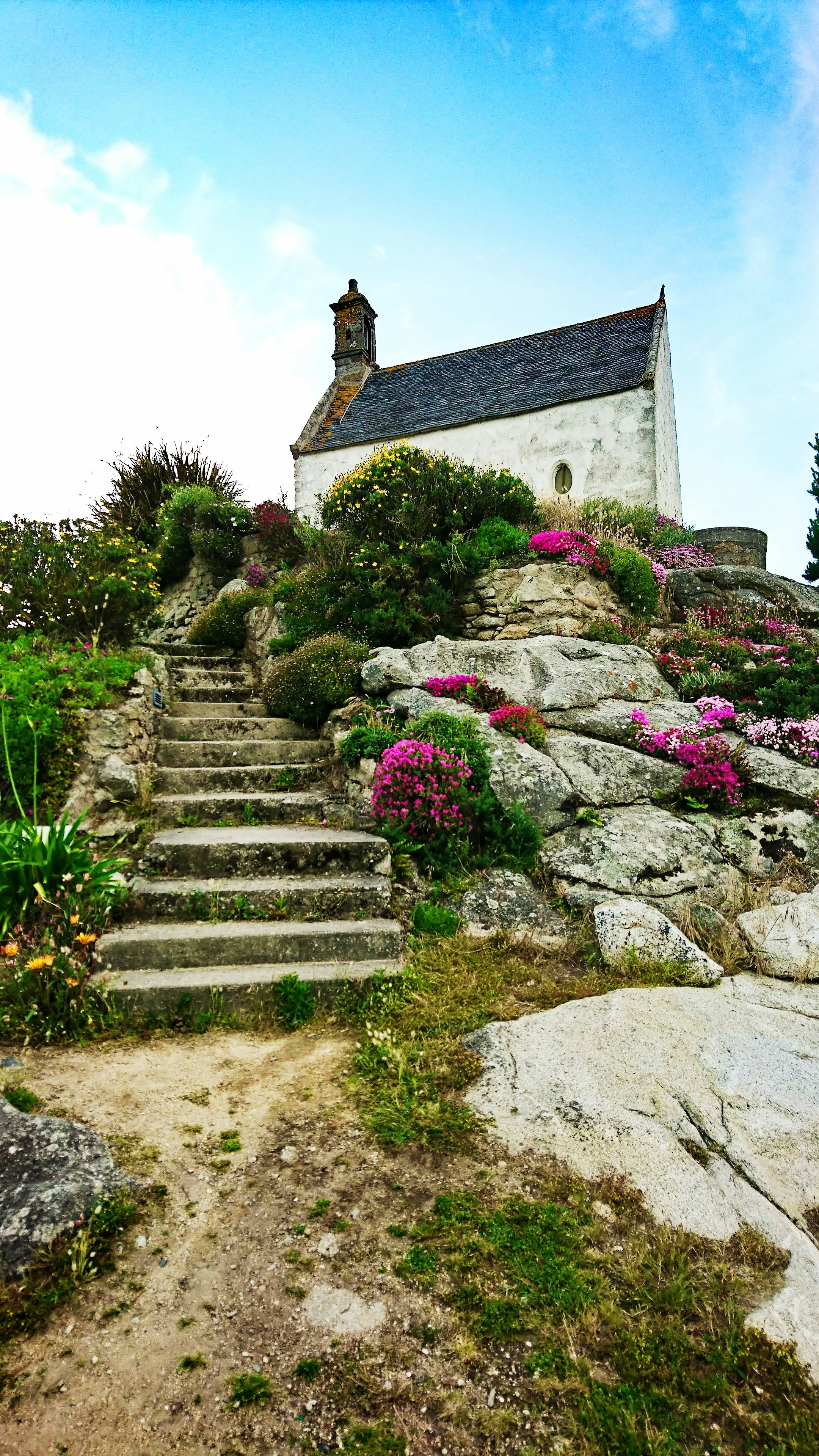 Charming stone chapel surrounded by vibrant flowers and rocky terrain, inviting visitors to explore its tranquil setting.