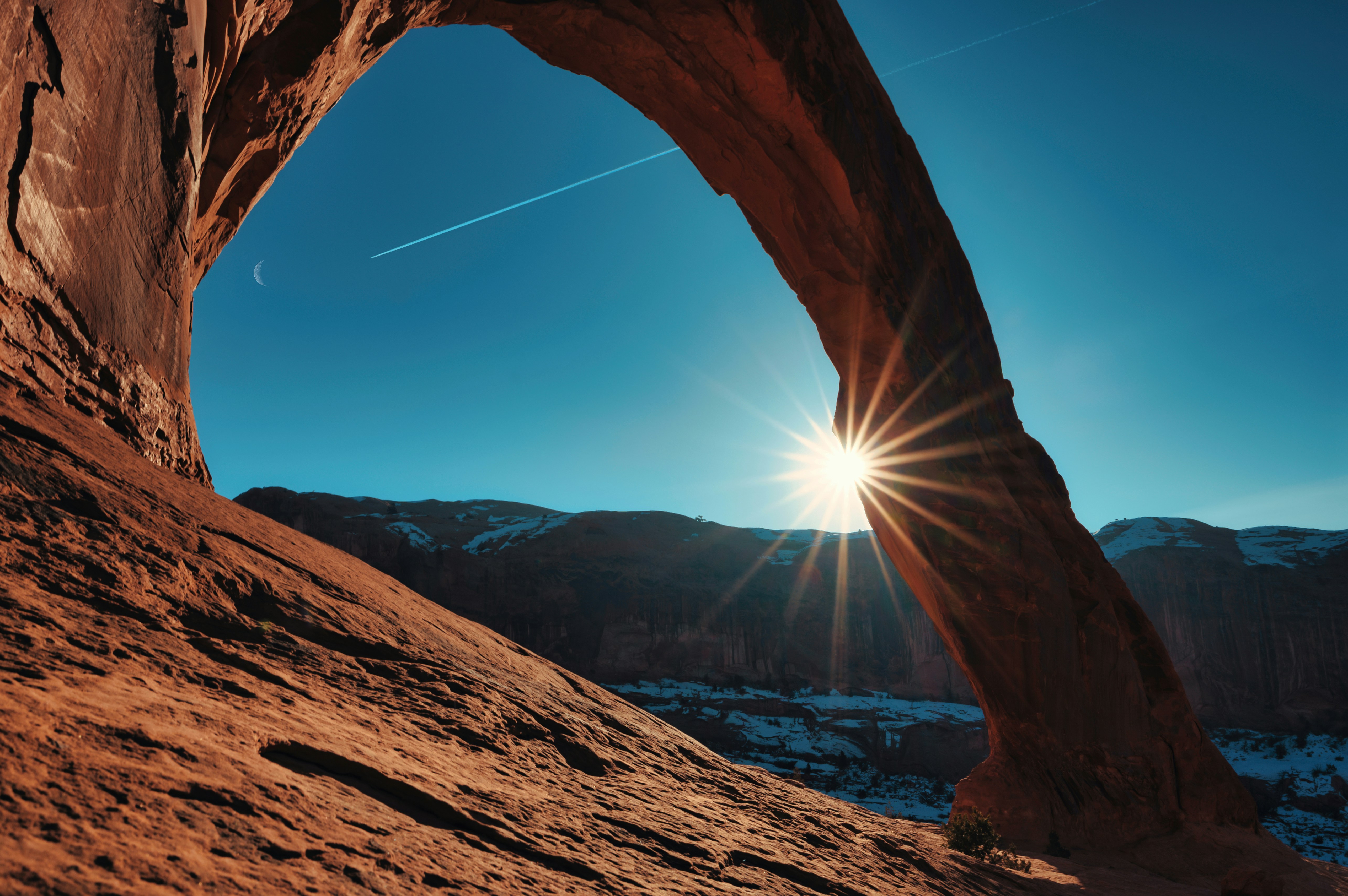 Natural arch framing the sun rising over a mountainous landscape, with a crescent moon visible in the sky.
