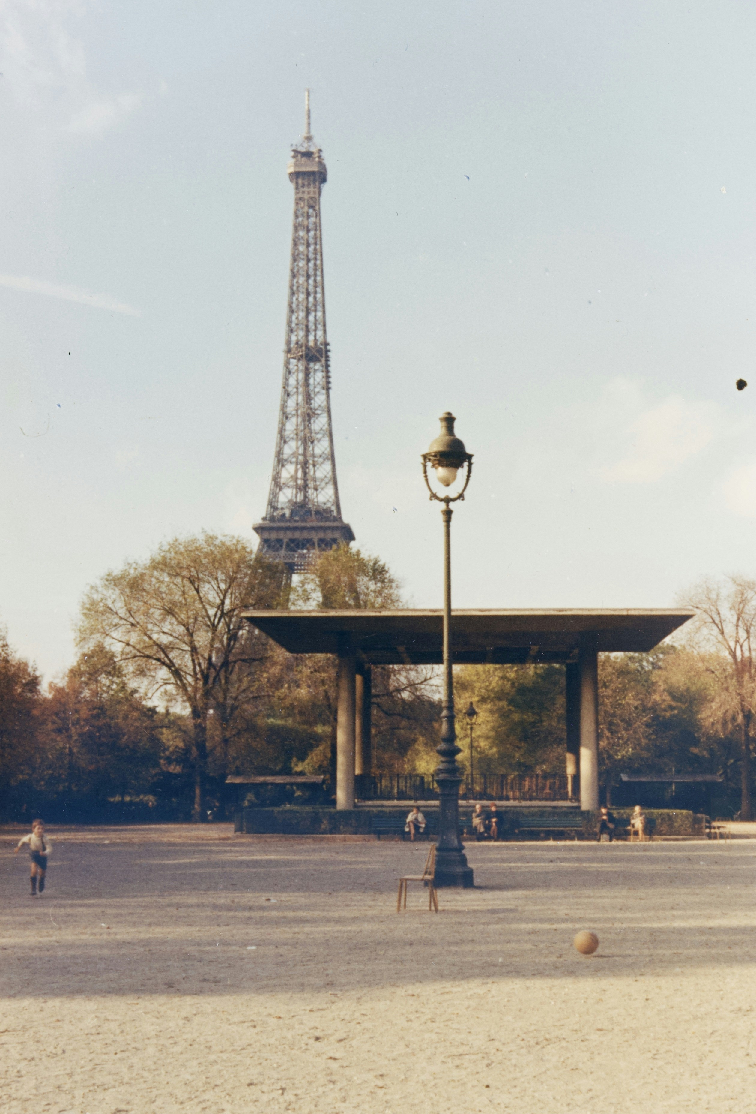 Eiffel Tower rises majestically behind a park pavilion, with a child playing in the foreground. The scene captures a serene moment in a Parisian park.