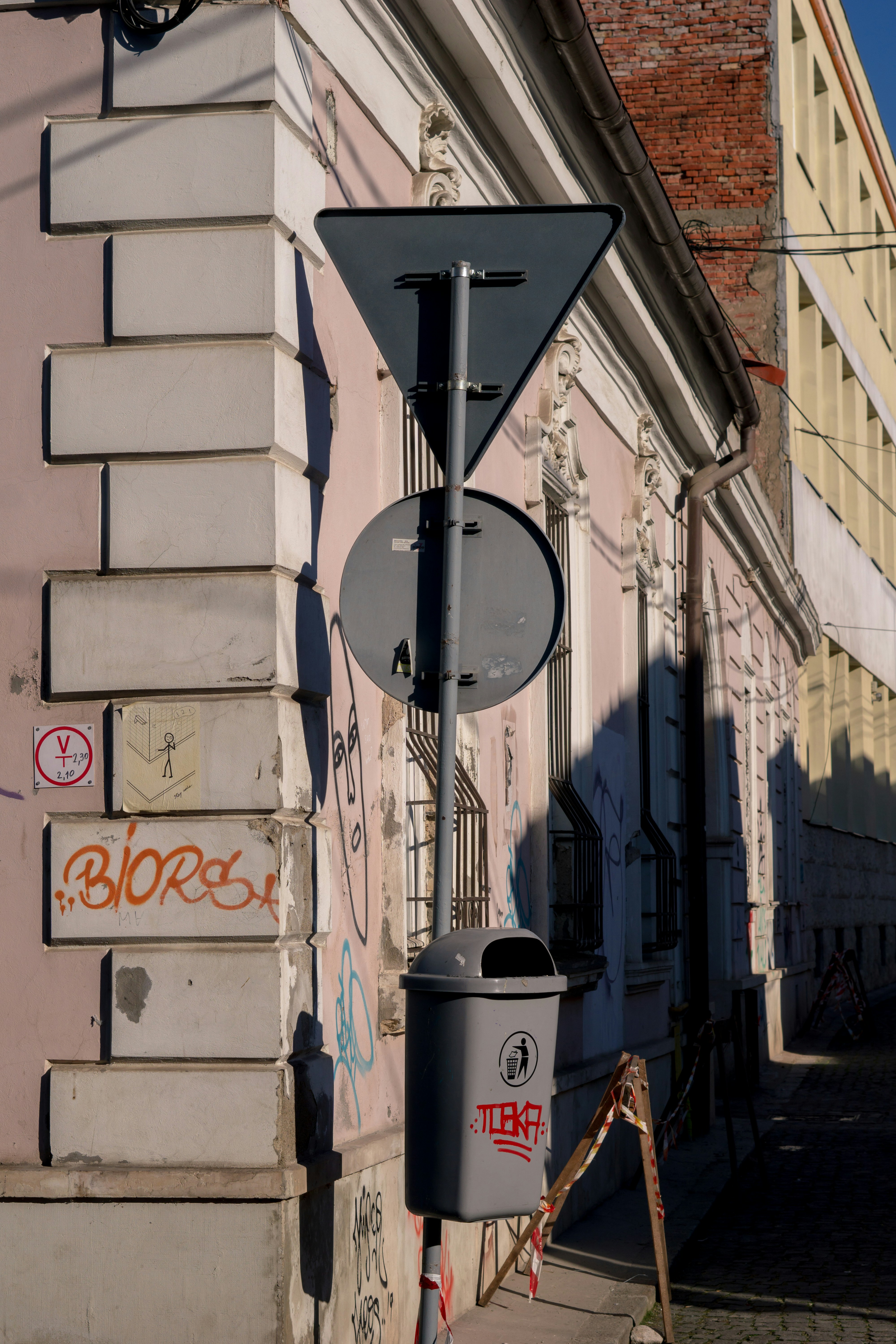 A triangular traffic sign and a circular road sign juxtaposed against a graffiti-covered wall in a sunlit urban setting.