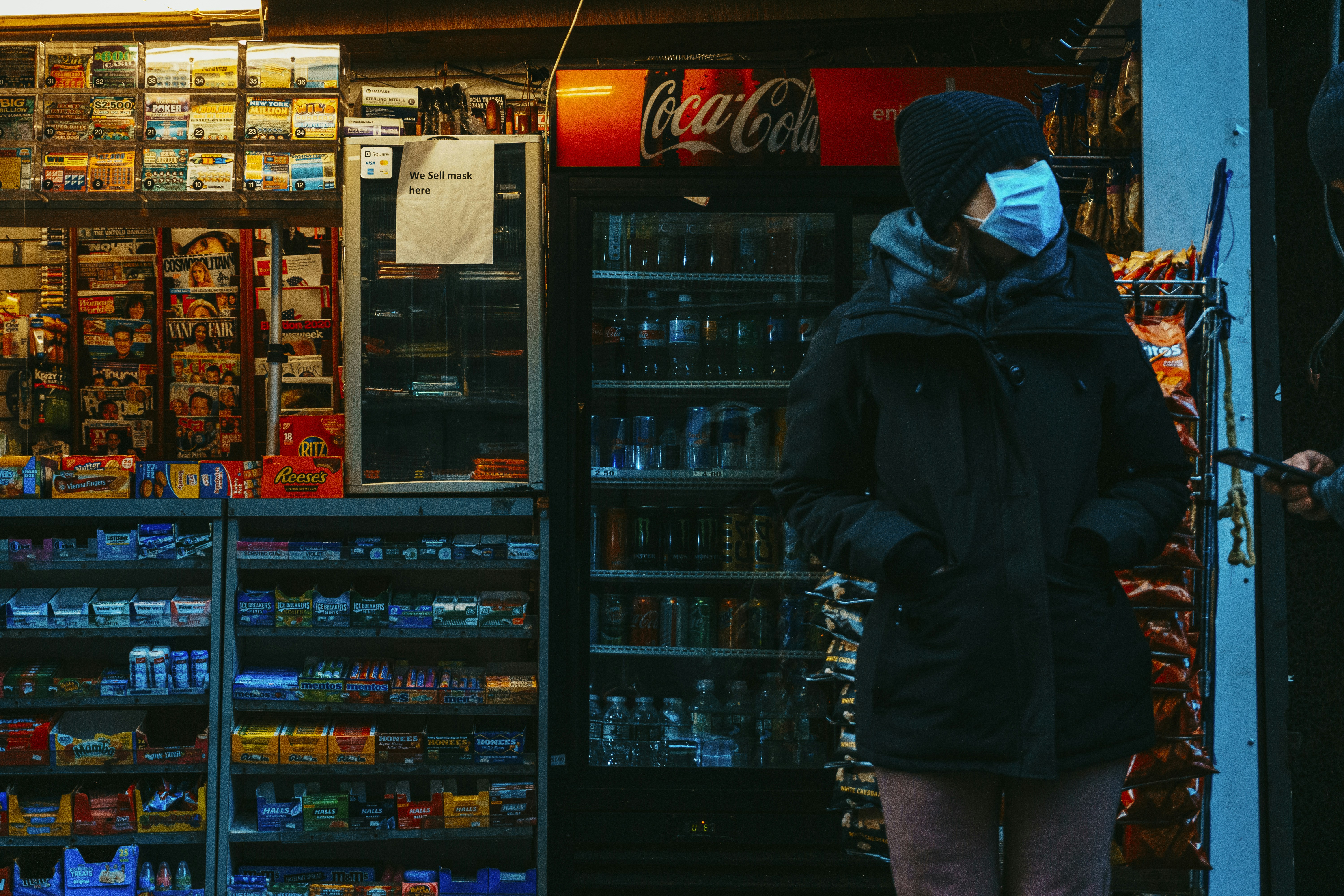 Person wearing a mask stands outside a convenience store in dim lighting.