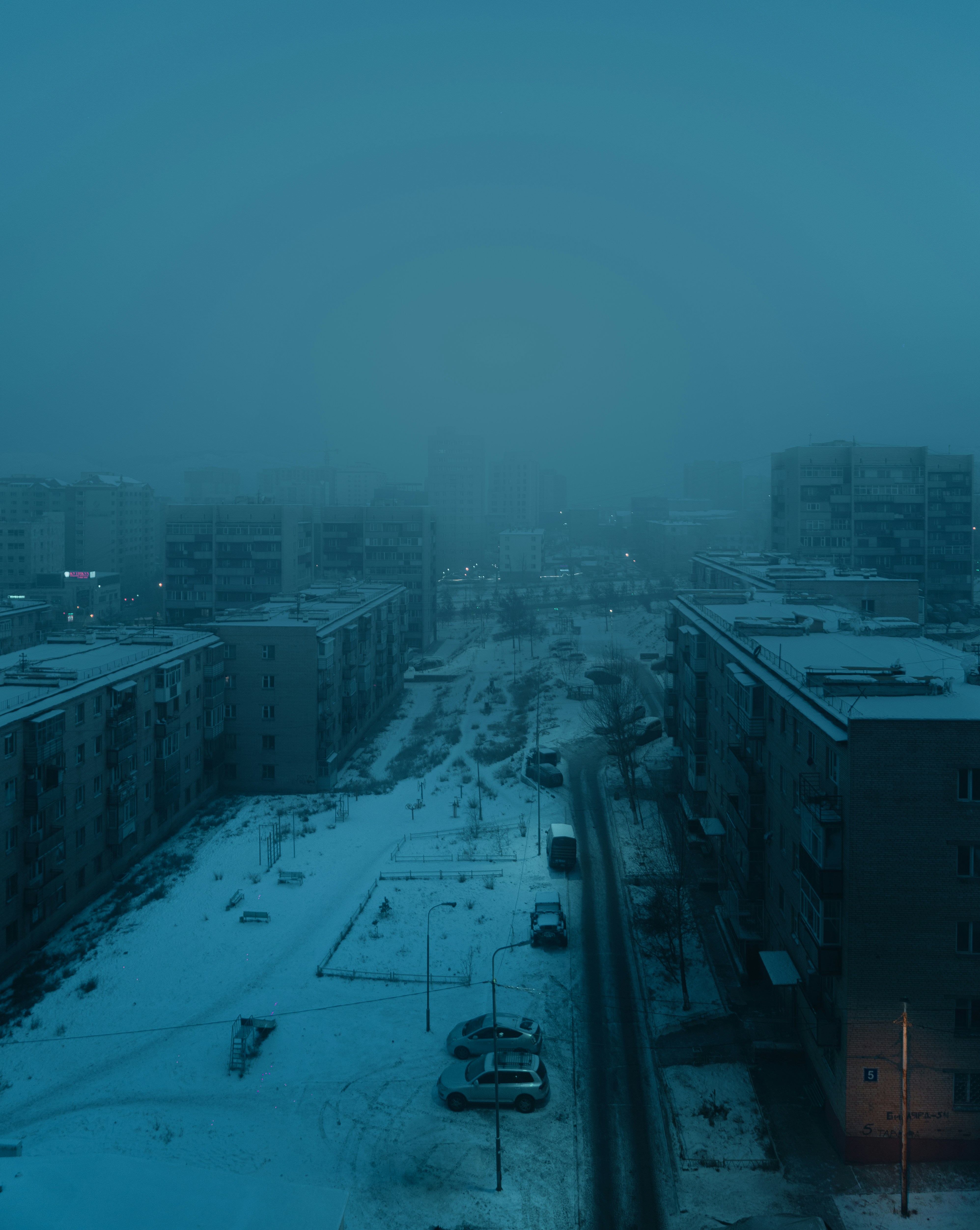 Snow-covered urban landscape shrouded in fog, showcasing apartment buildings and a quiet street. The scene conveys a serene yet eerie atmosphere.
