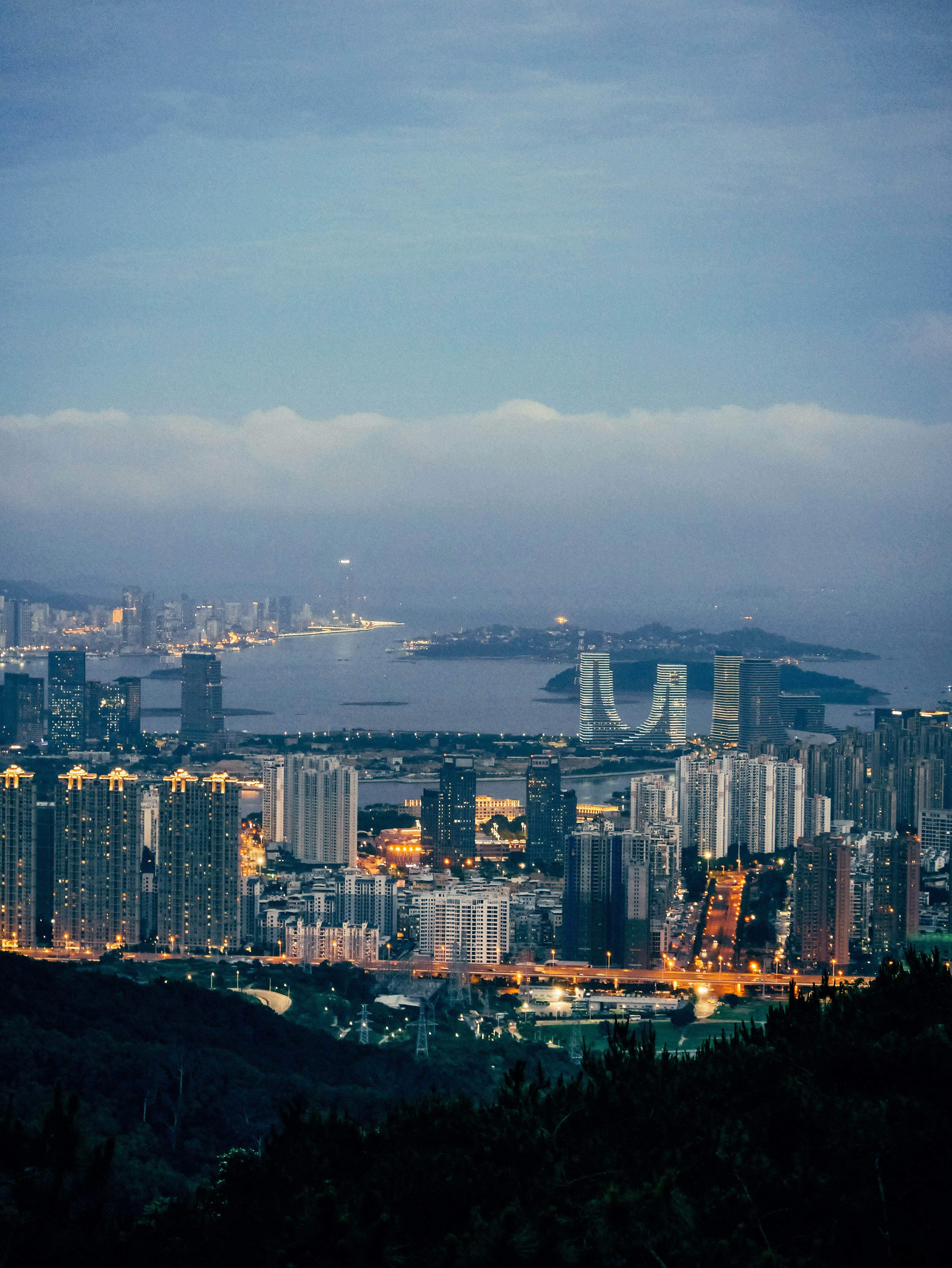 A panoramic view of a city skyline at twilight, showcasing illuminated buildings and a serene harbor under a cloudy sky.