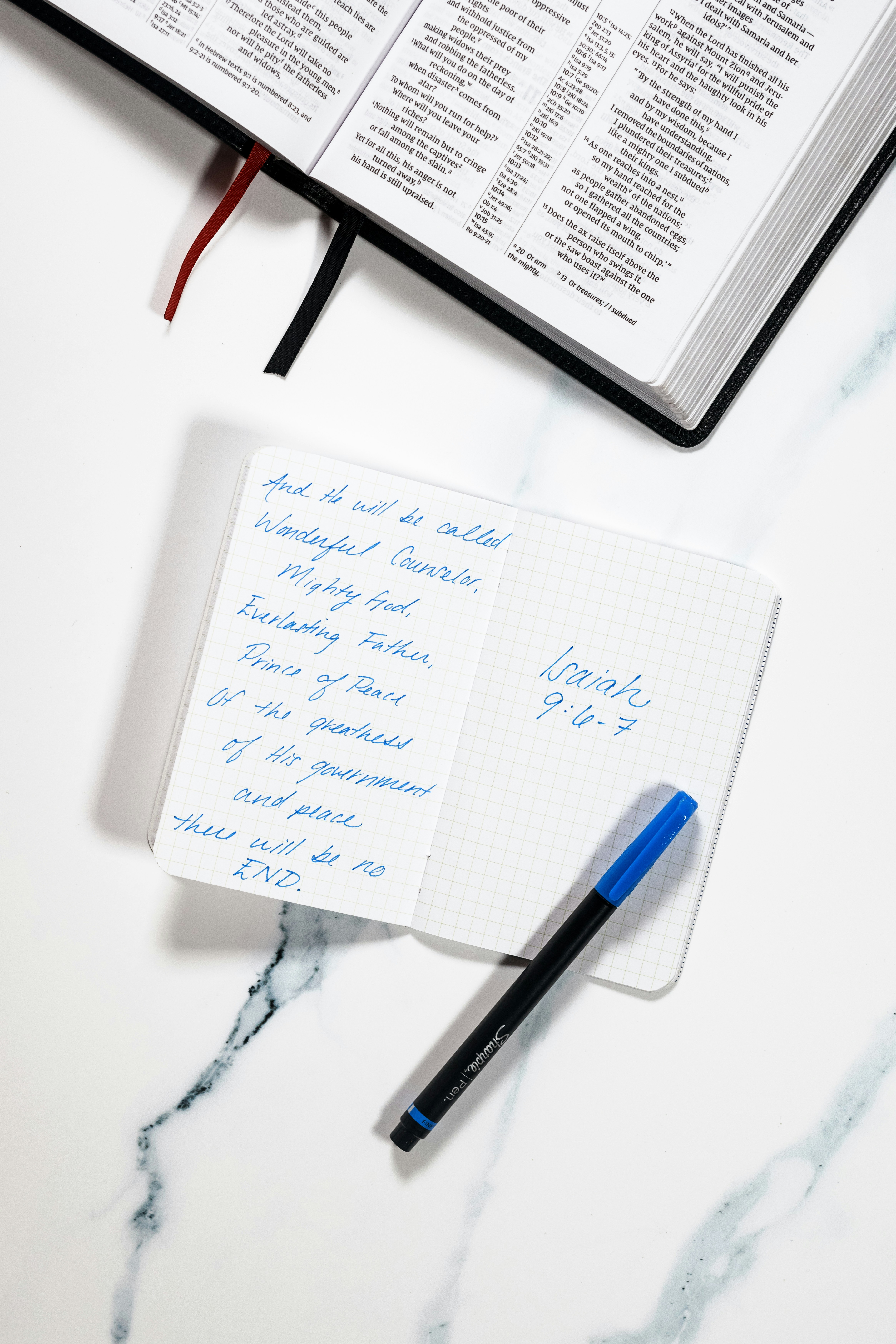 Open notebook with handwritten notes and a blue pen resting on a marble surface, alongside an open Bible. The text references Isaiah 9:6-7.