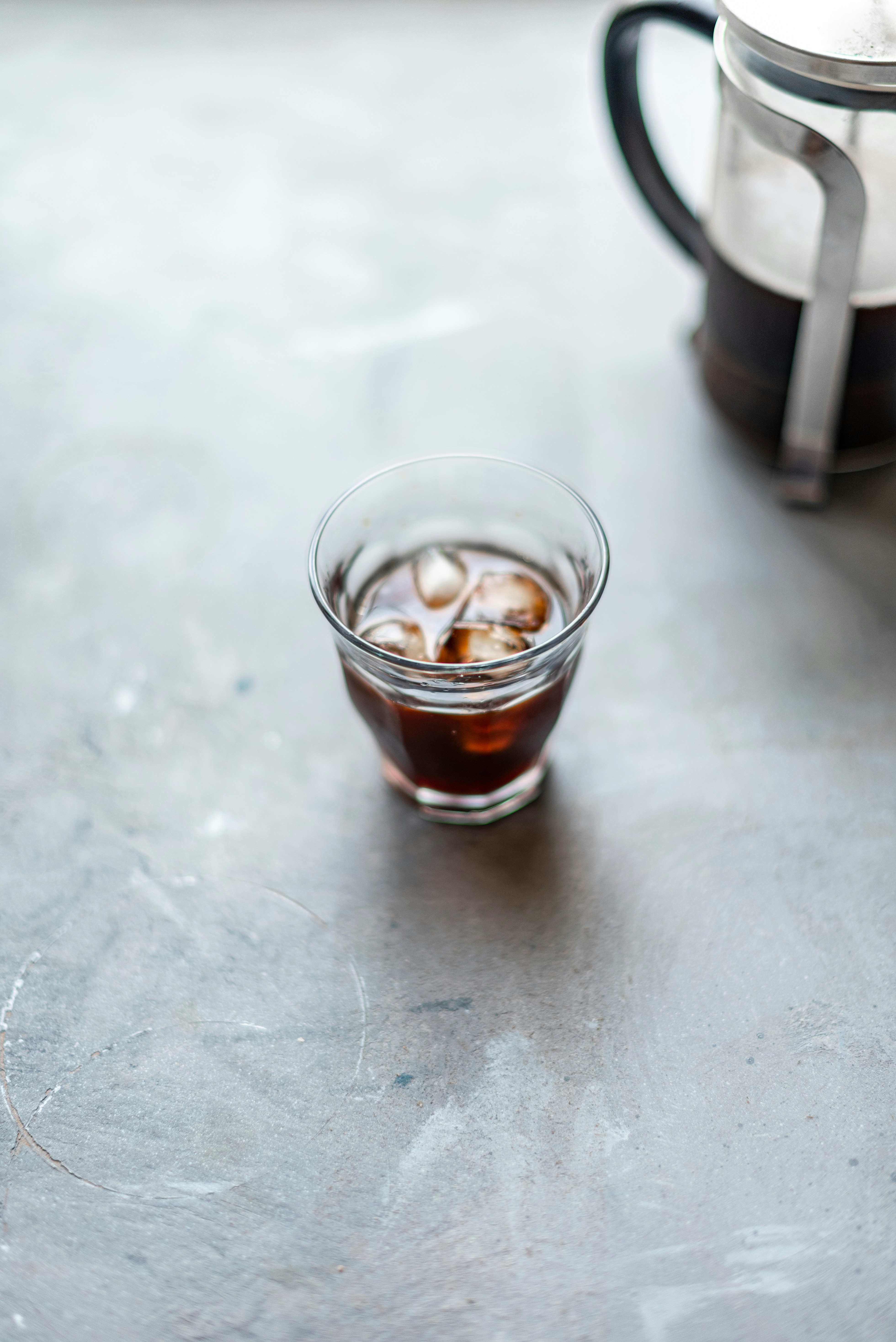 Glass of iced coffee with ice cubes on a textured gray surface, accompanied by a French press in the background.