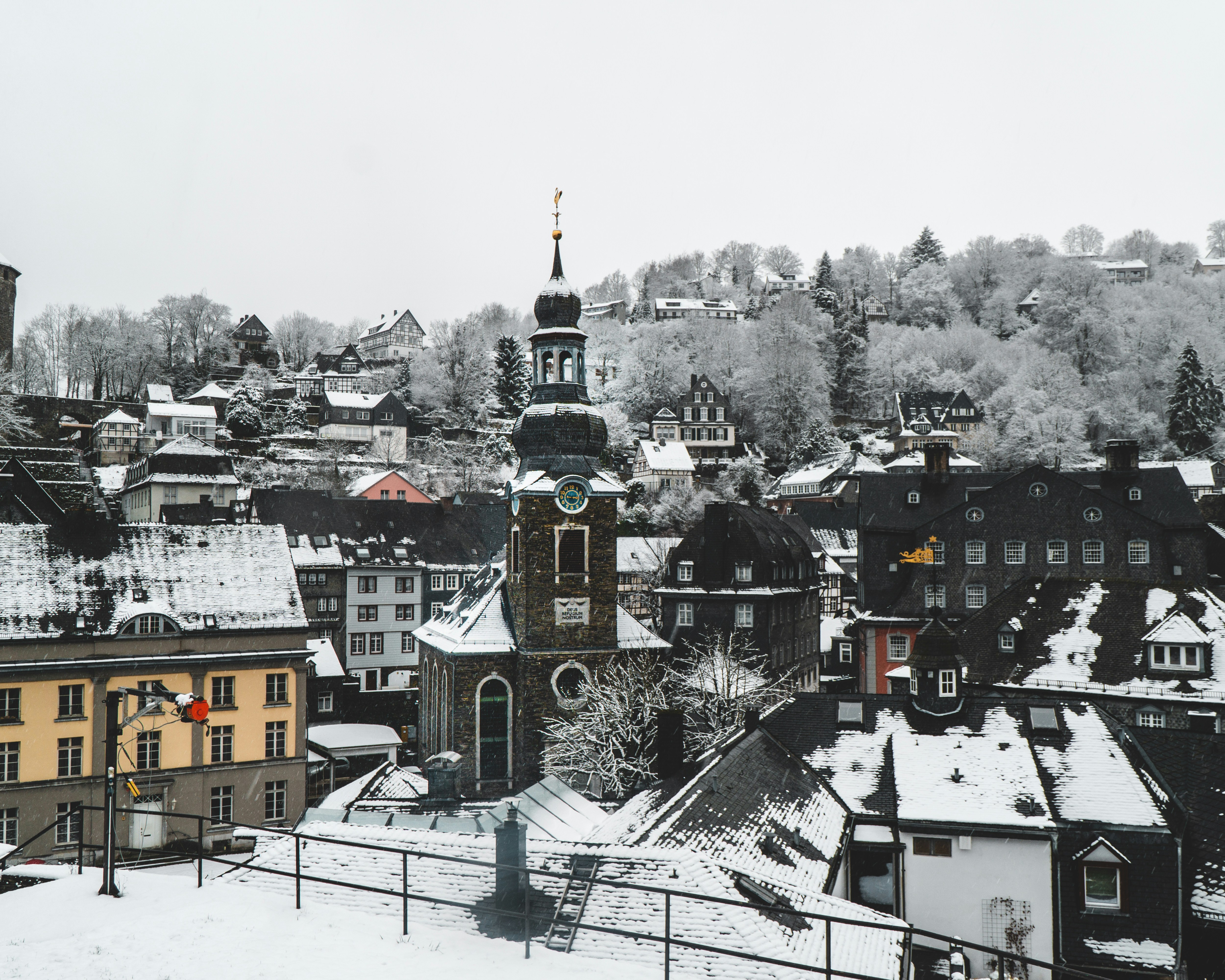 Charming village blanketed in snow, featuring a prominent clock tower amidst quaint houses and frosted trees. A serene winter scene unfolds in the background.