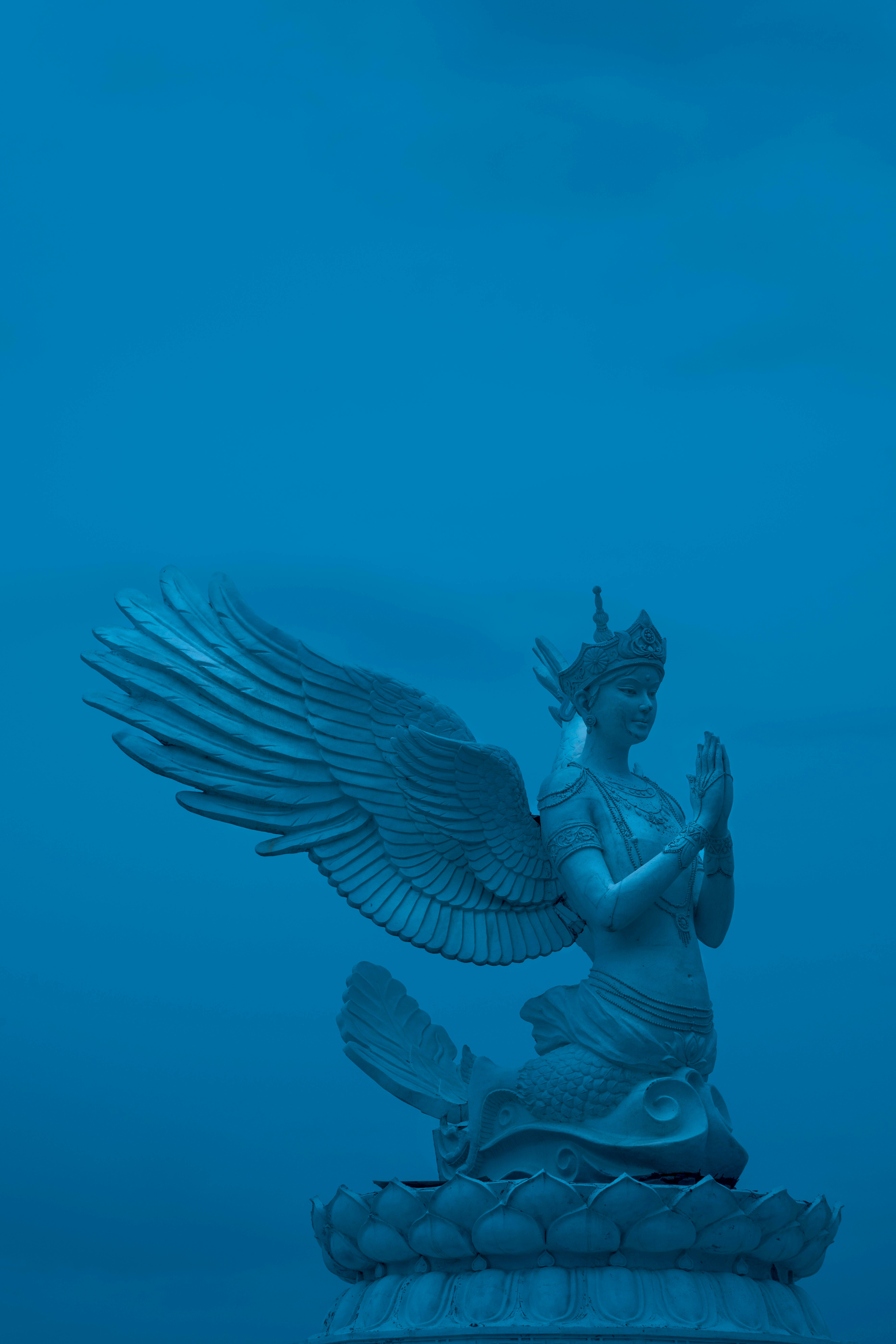 Ornate winged statue in prayer against a vivid blue sky.