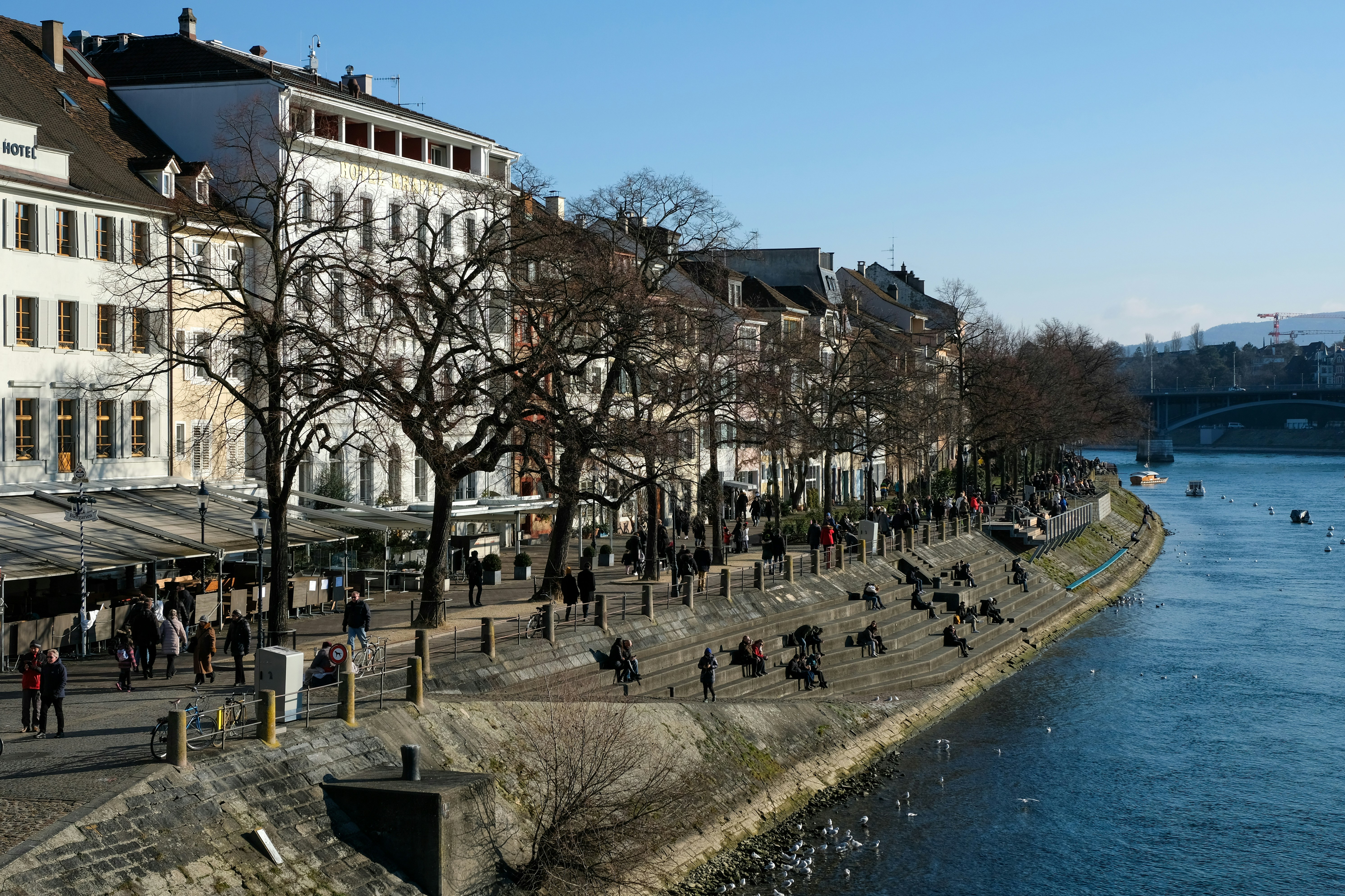 People walking along a tree-lined sidewalk beside a tranquil river under a clear blue sky.