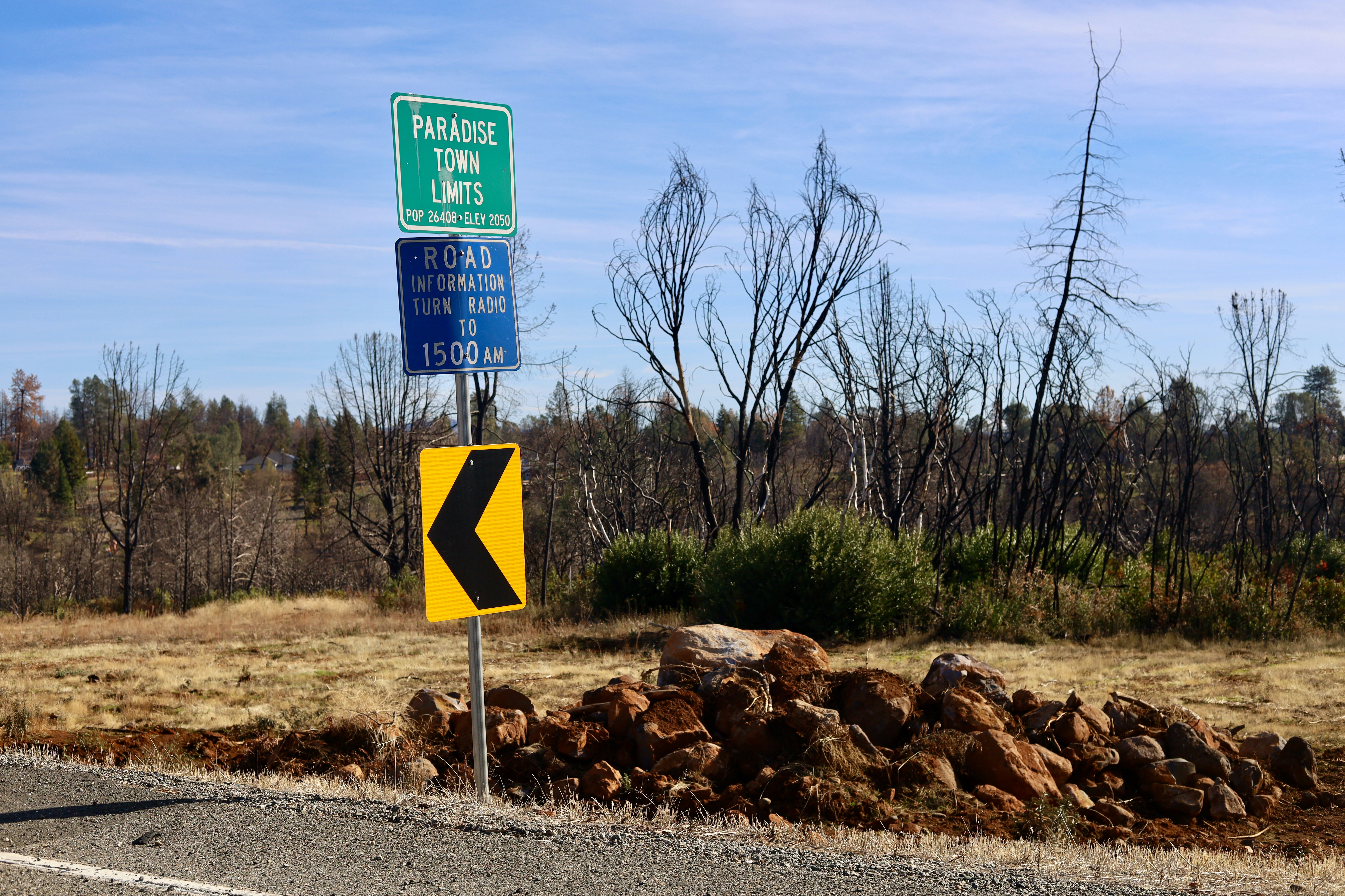 Road sign indicating the limits of Paradise town, with a warning for a curve ahead, set against a backdrop of recovering landscape.