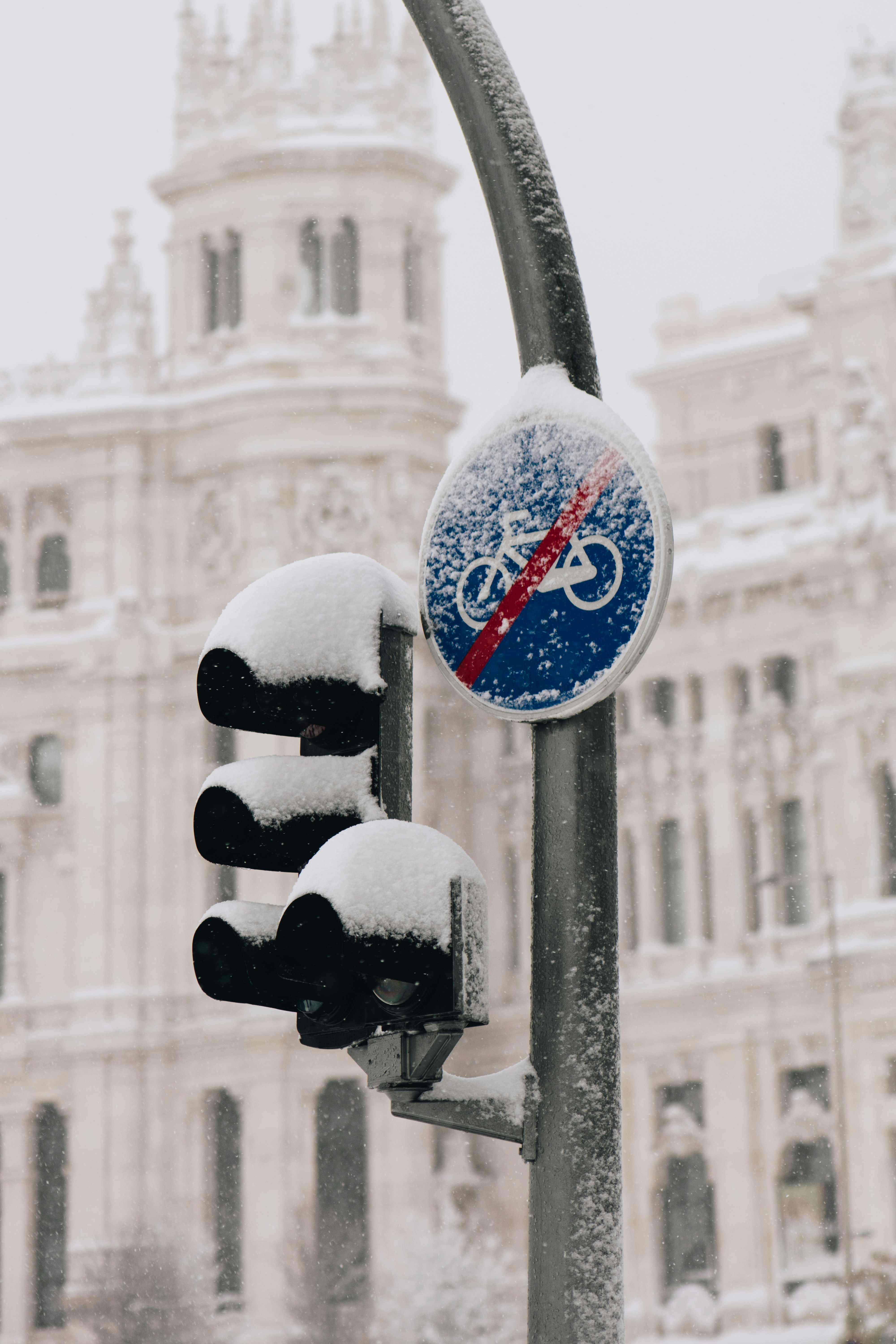 Snow-covered traffic lights and a bicycle prohibition sign stand in stark contrast against a historic building backdrop during a winter snowstorm.