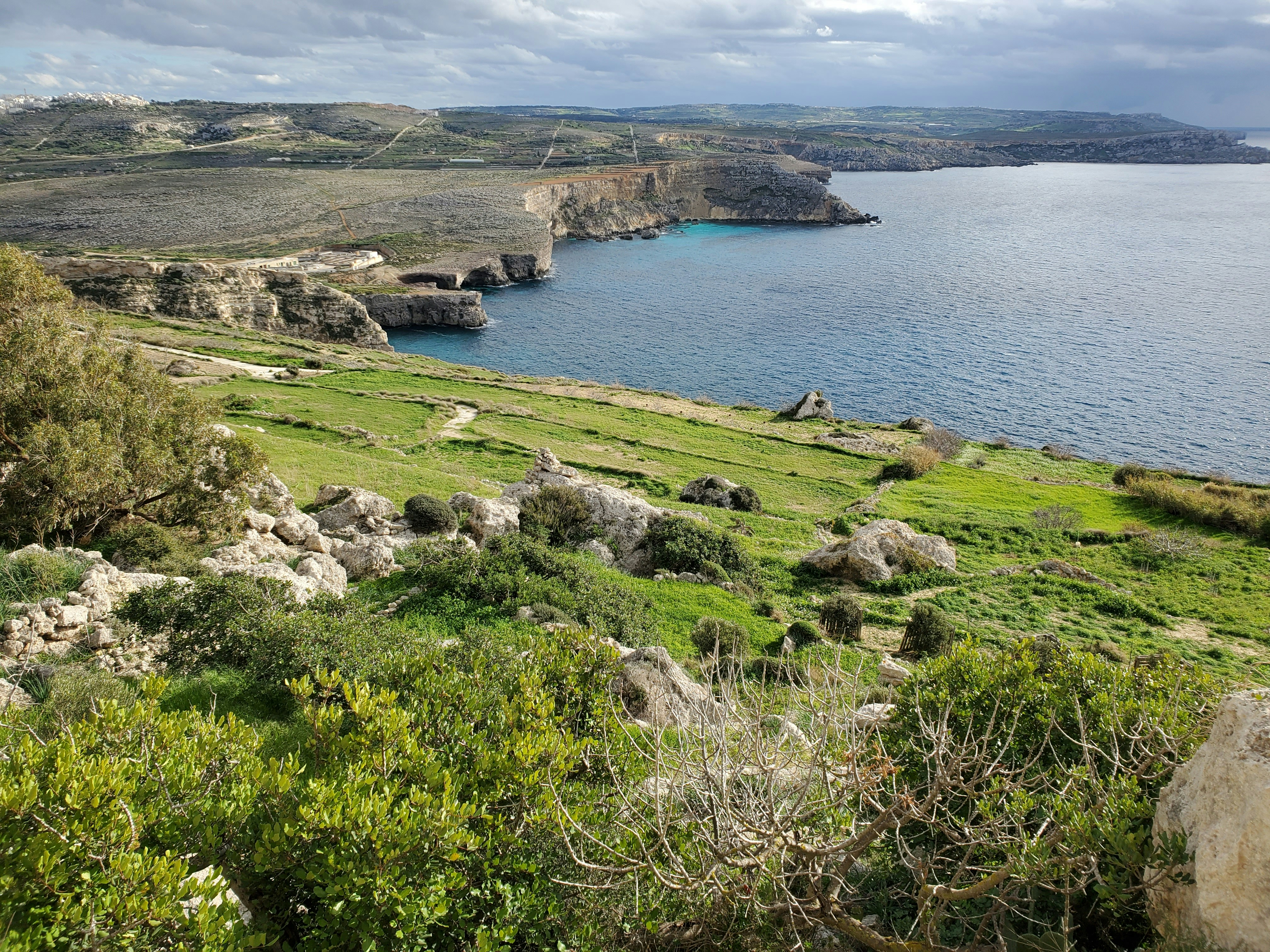 Lush green landscape cascading down to rocky cliffs by the tranquil sea, with a dramatic sky overhead.
