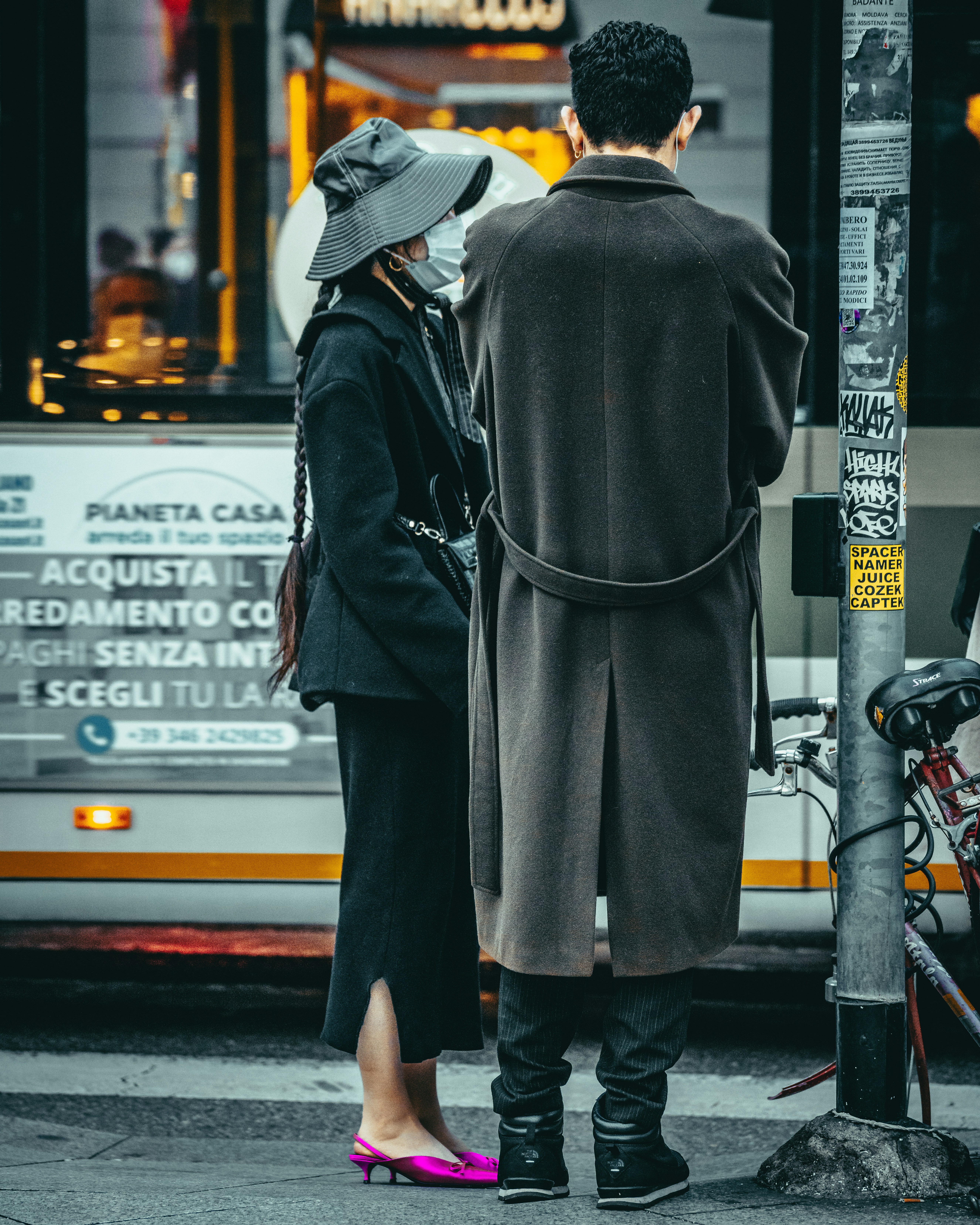 Two stylish individuals engage in conversation on a bustling street, framed by urban elements and soft city lights.
