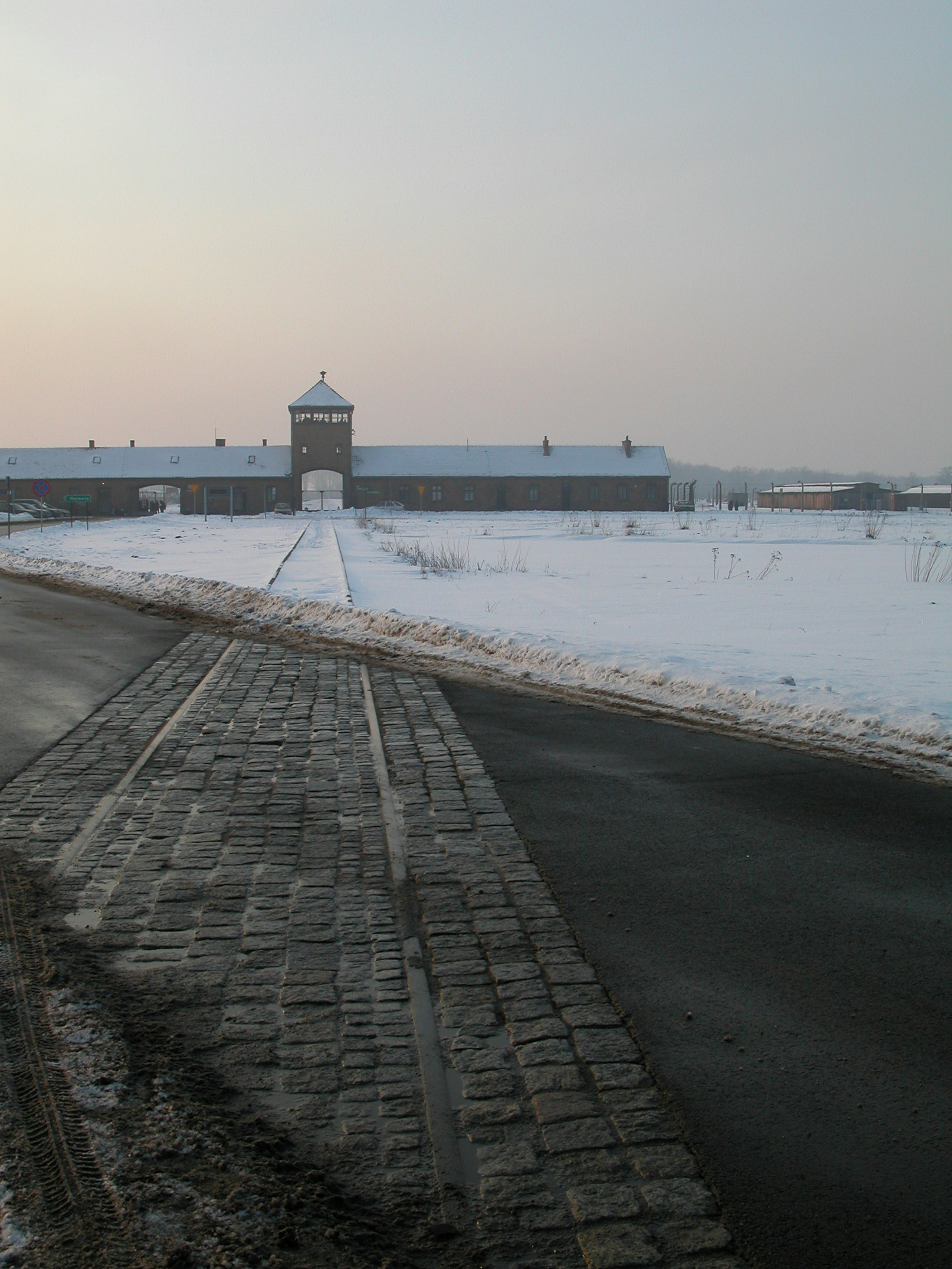 Snow-covered pathway leading to a historical site, framed by a muted sky at dawn. The architecture hints at a somber past.