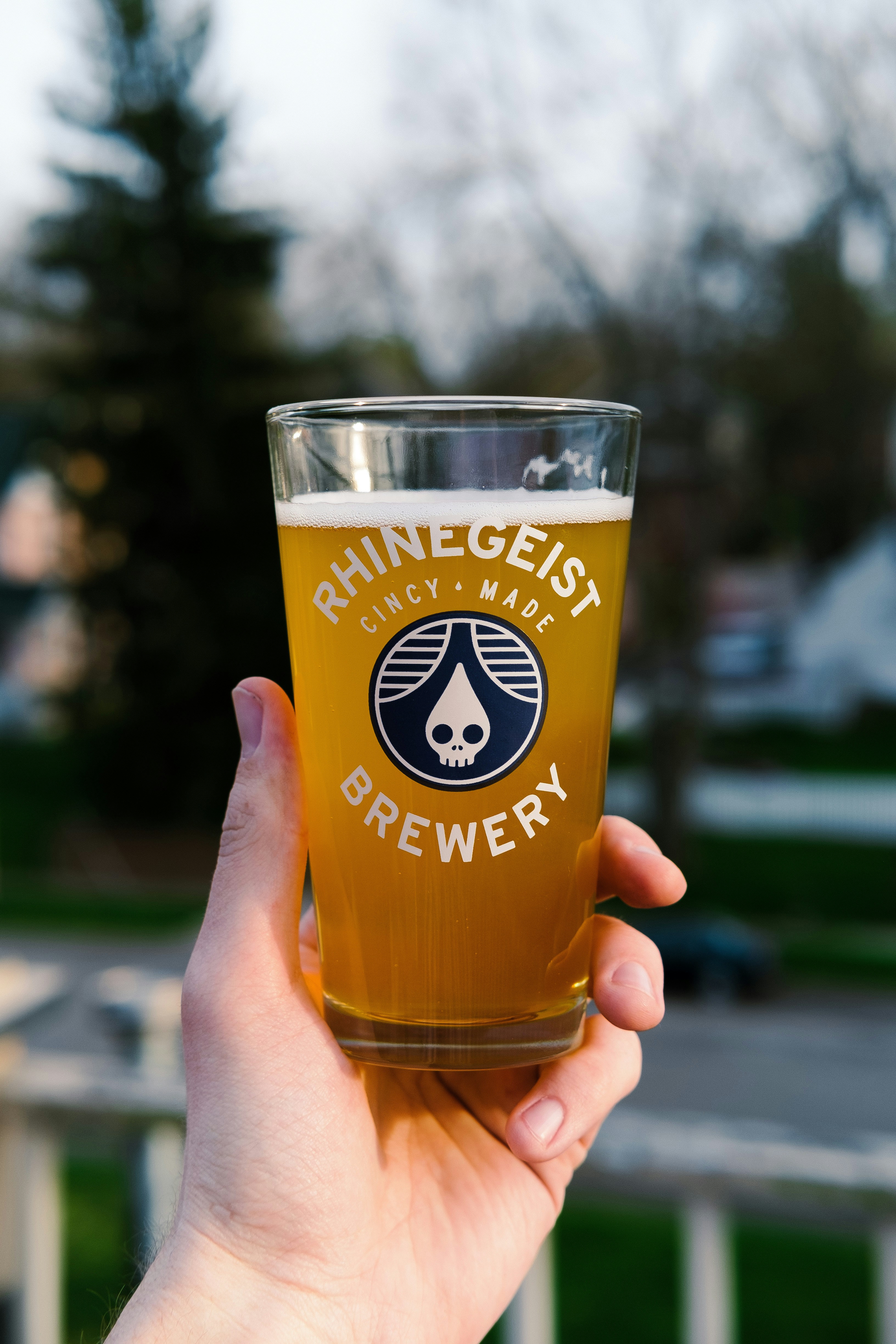 Hand holding a glass of Rhinegeist Brewery beer, showcasing the logo prominently against a blurred outdoor backdrop.