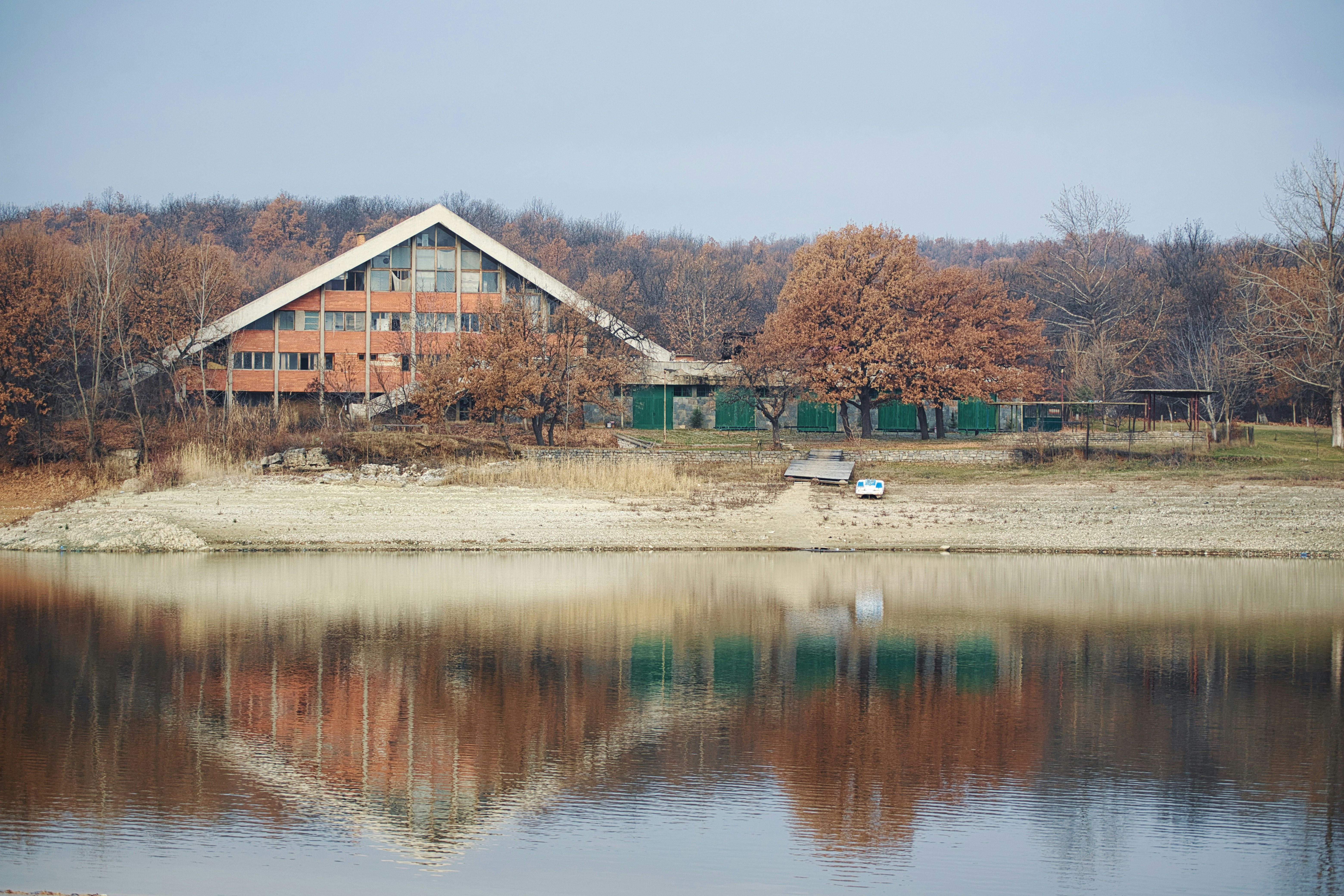 Lodge with a triangular roof reflected in a calm lake, surrounded by autumn trees.