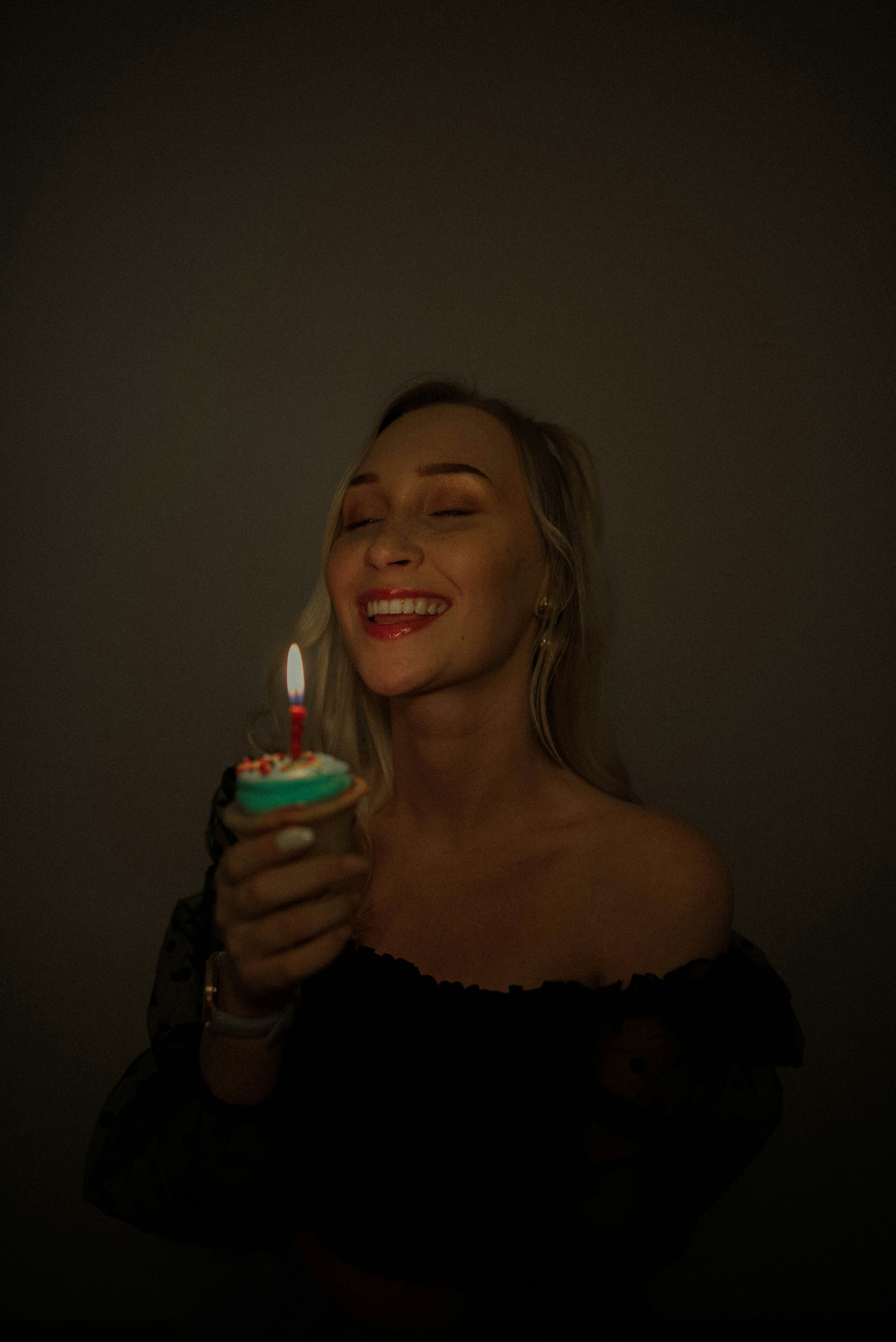 Woman with a joyful expression holding a cupcake with a lit candle against a dark background.