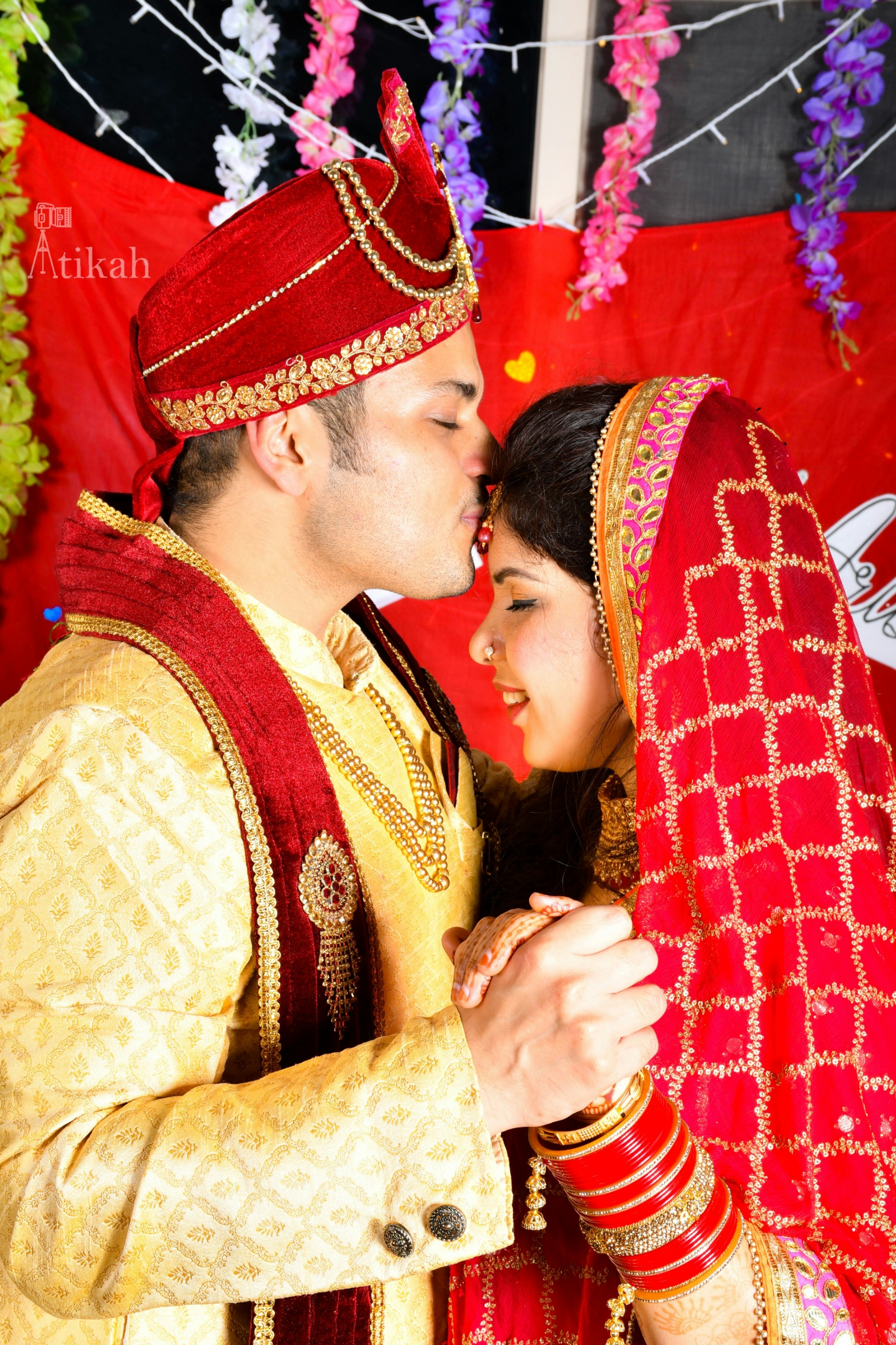 Crimson Veil, Golden Hands: A Nuptial Kiss Amid Floral Garlands