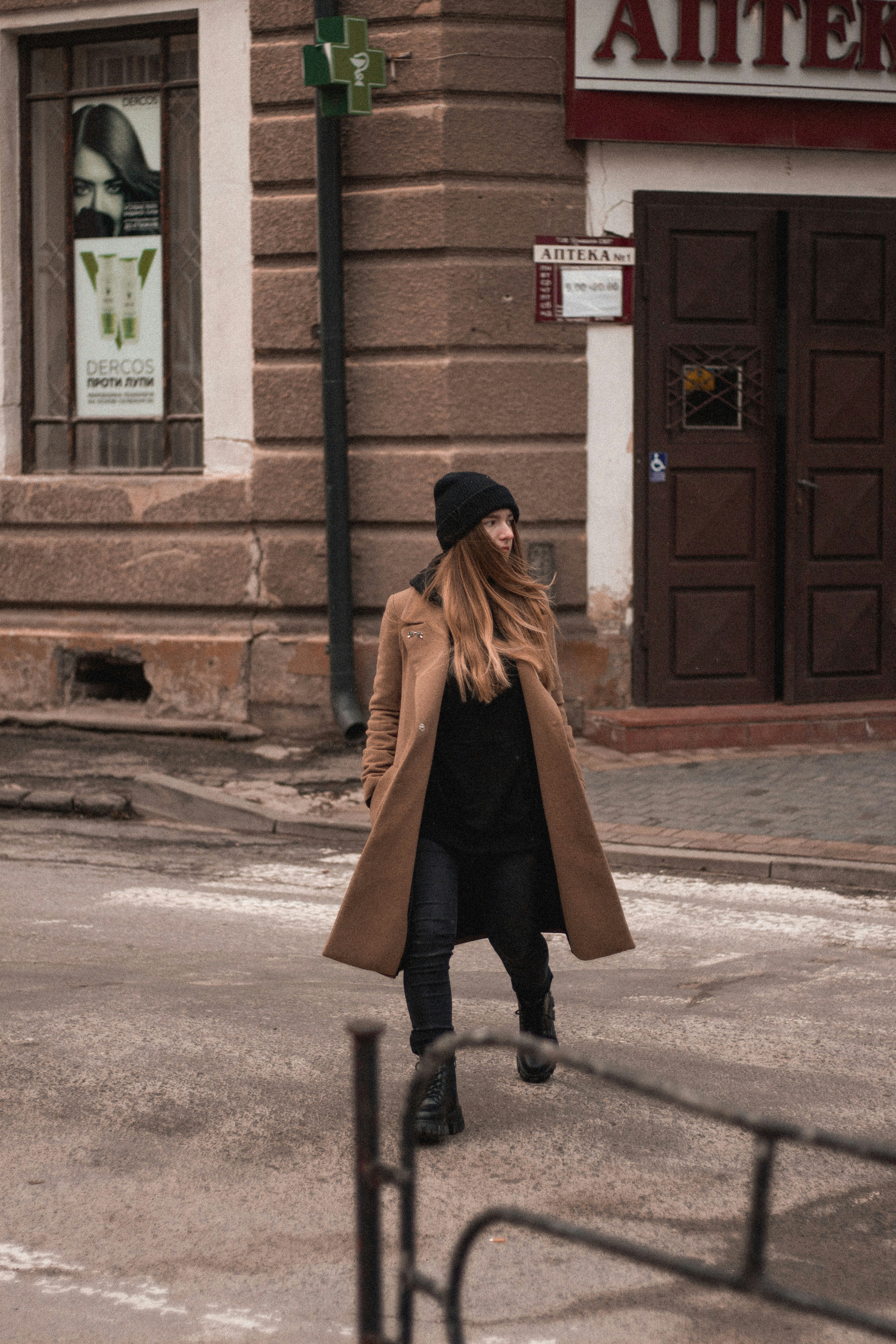 A woman in a stylish coat walks across a city street, framed by a vintage building and a pharmacy sign. The scene captures the essence of urban life.