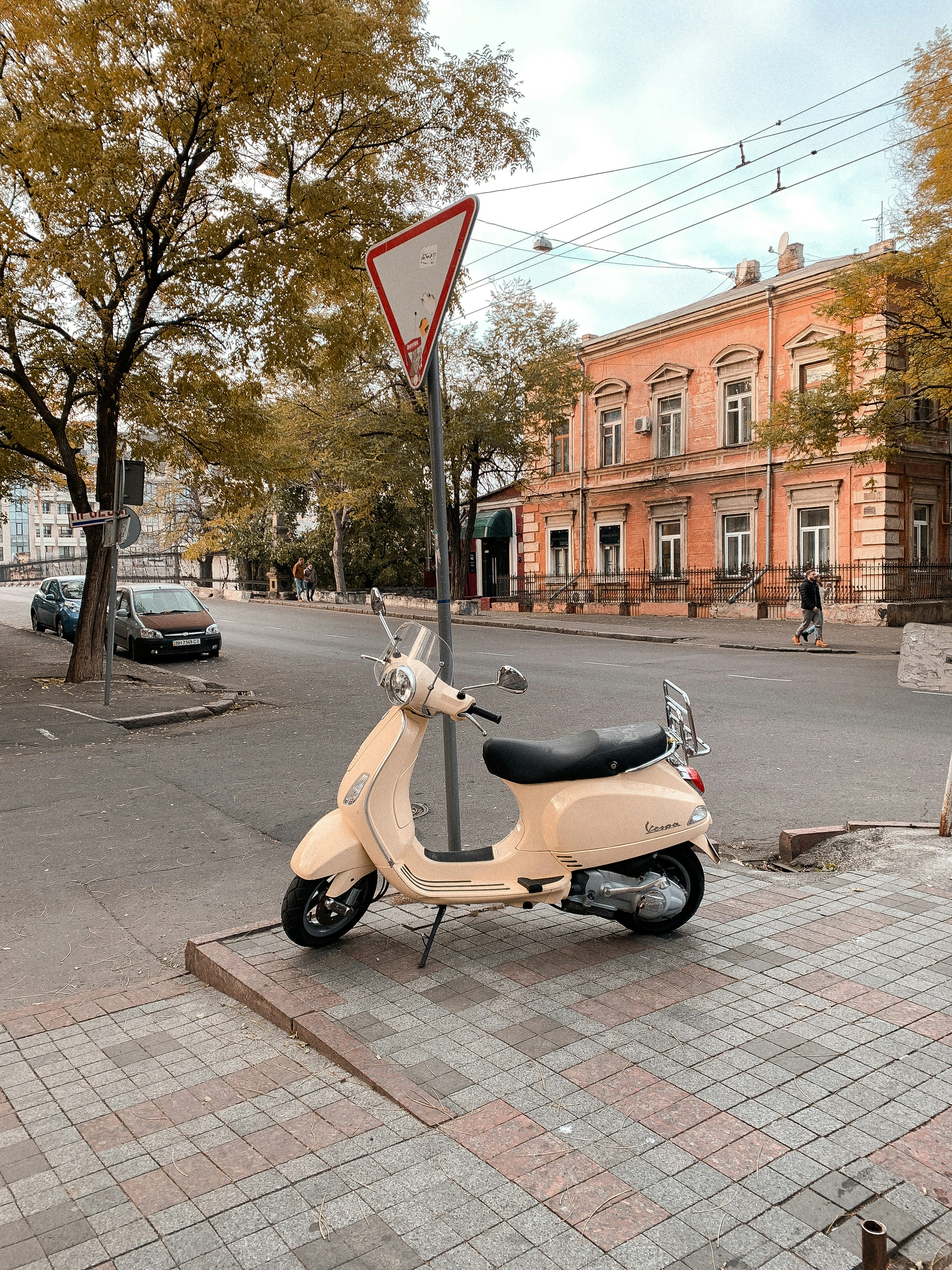 A vintage scooter parked on a city street, framed by autumn foliage and historic architecture. The scene captures a tranquil urban atmosphere.