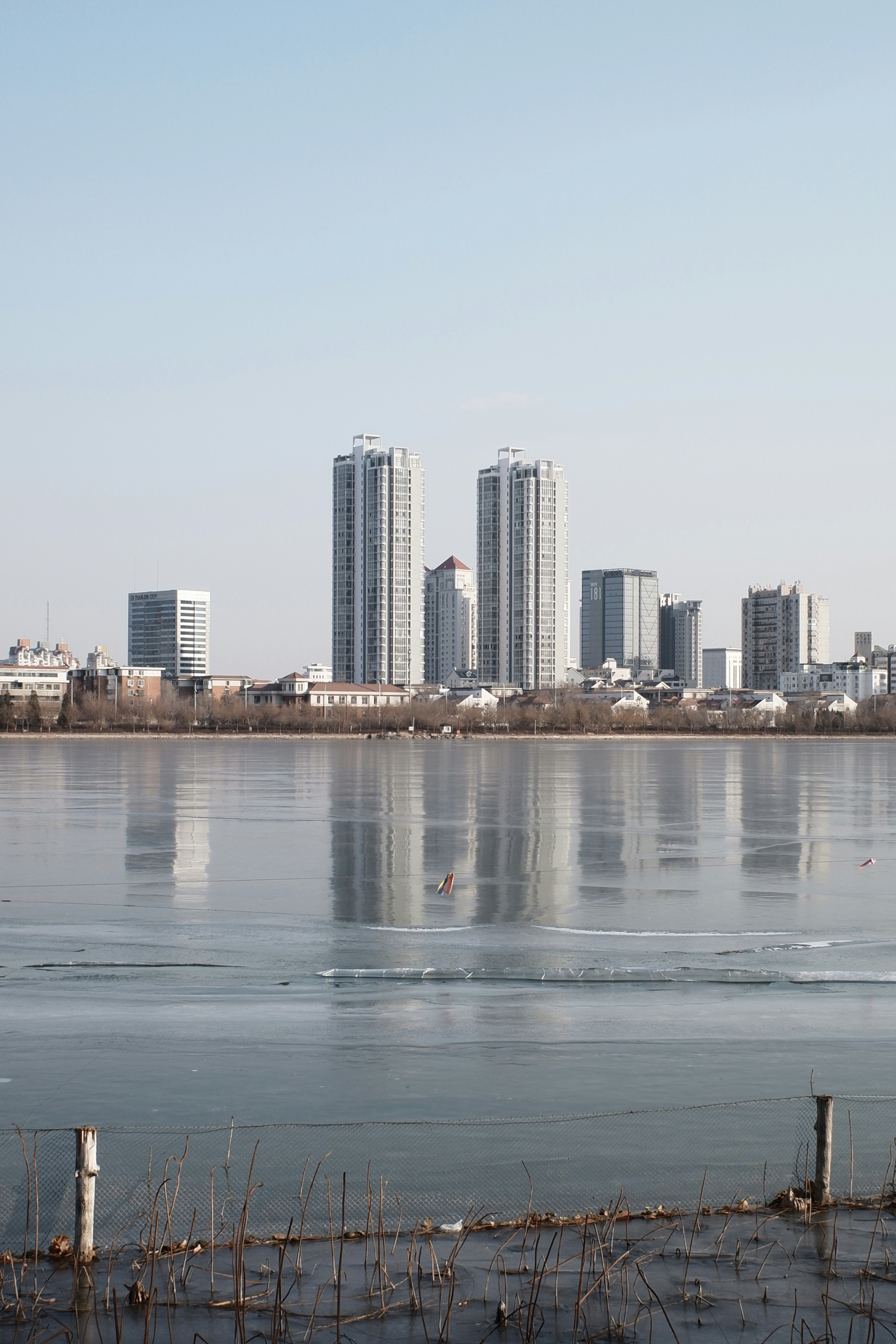 Modern skyscrapers reflecting on a frozen lake, framed by sparse vegetation in the foreground. The scene captures the serene juxtaposition of nature and urban life.