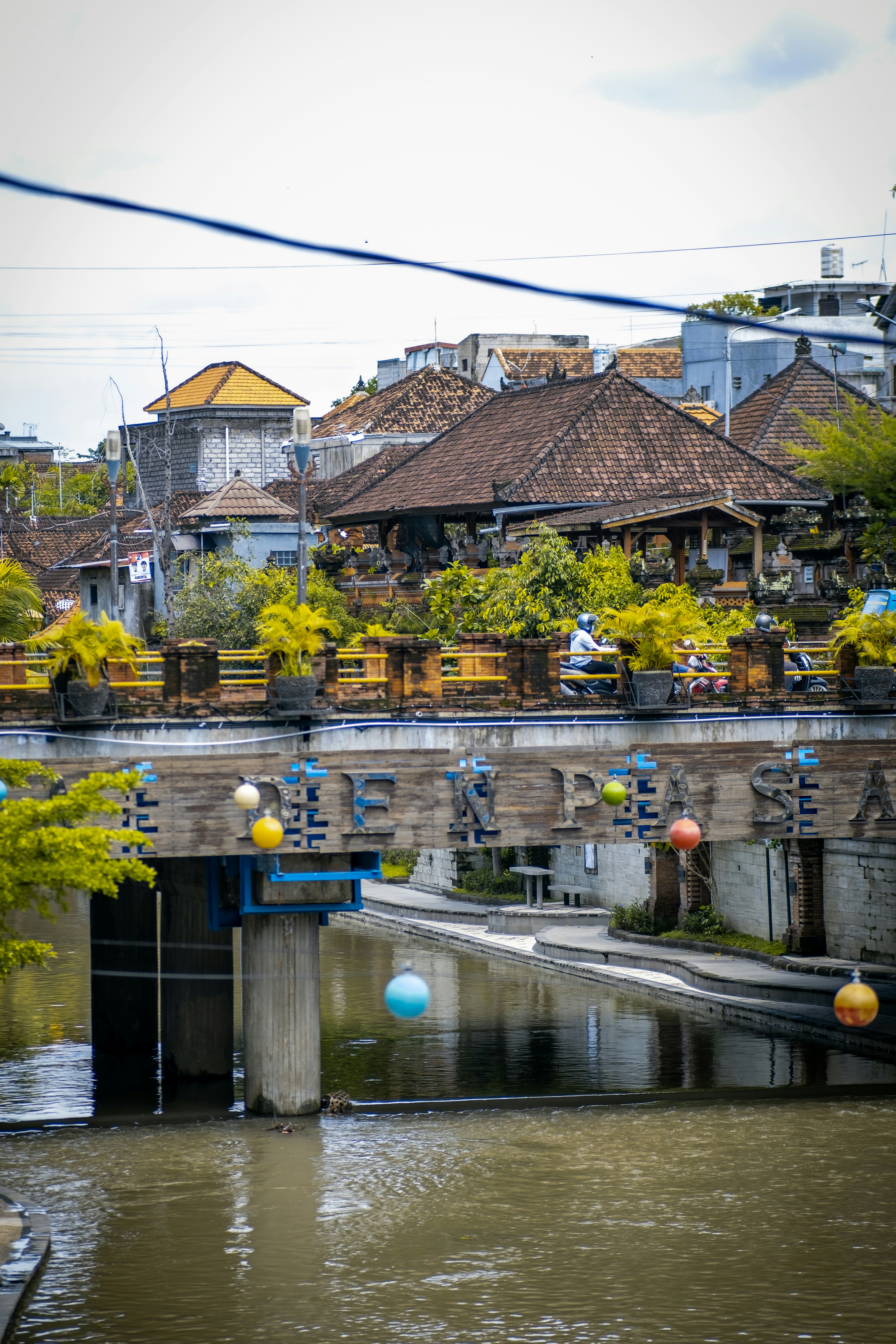 Wooden bridge adorned with colorful lanterns spans over a tranquil river, framed by lush greenery and rustic rooftops.