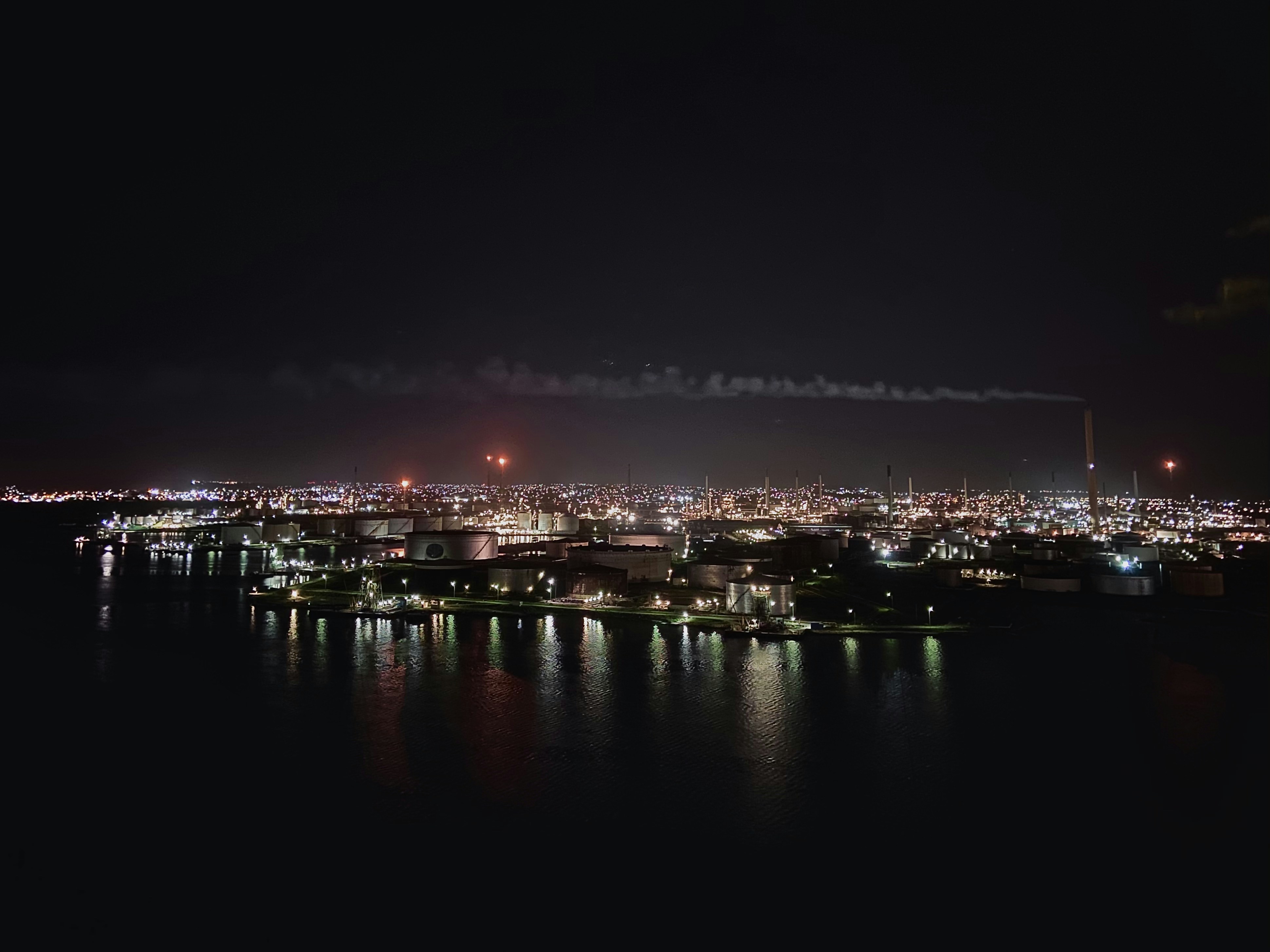 Illuminated industrial landscape reflecting on water at night, showcasing a network of tanks and lights.