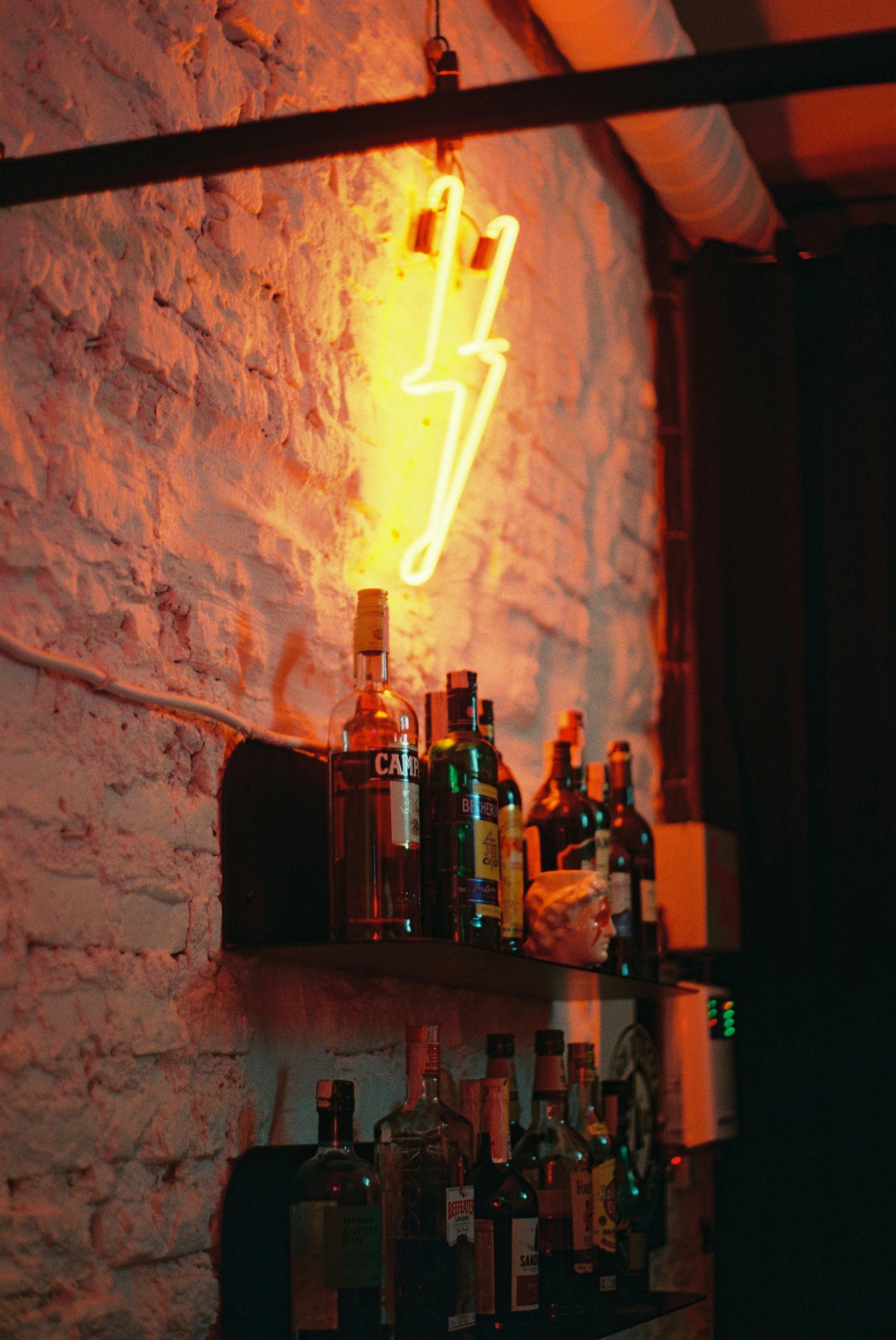 Neon lightning sign glows above a shelf of liquor bottles mounted on a rough brick wall.