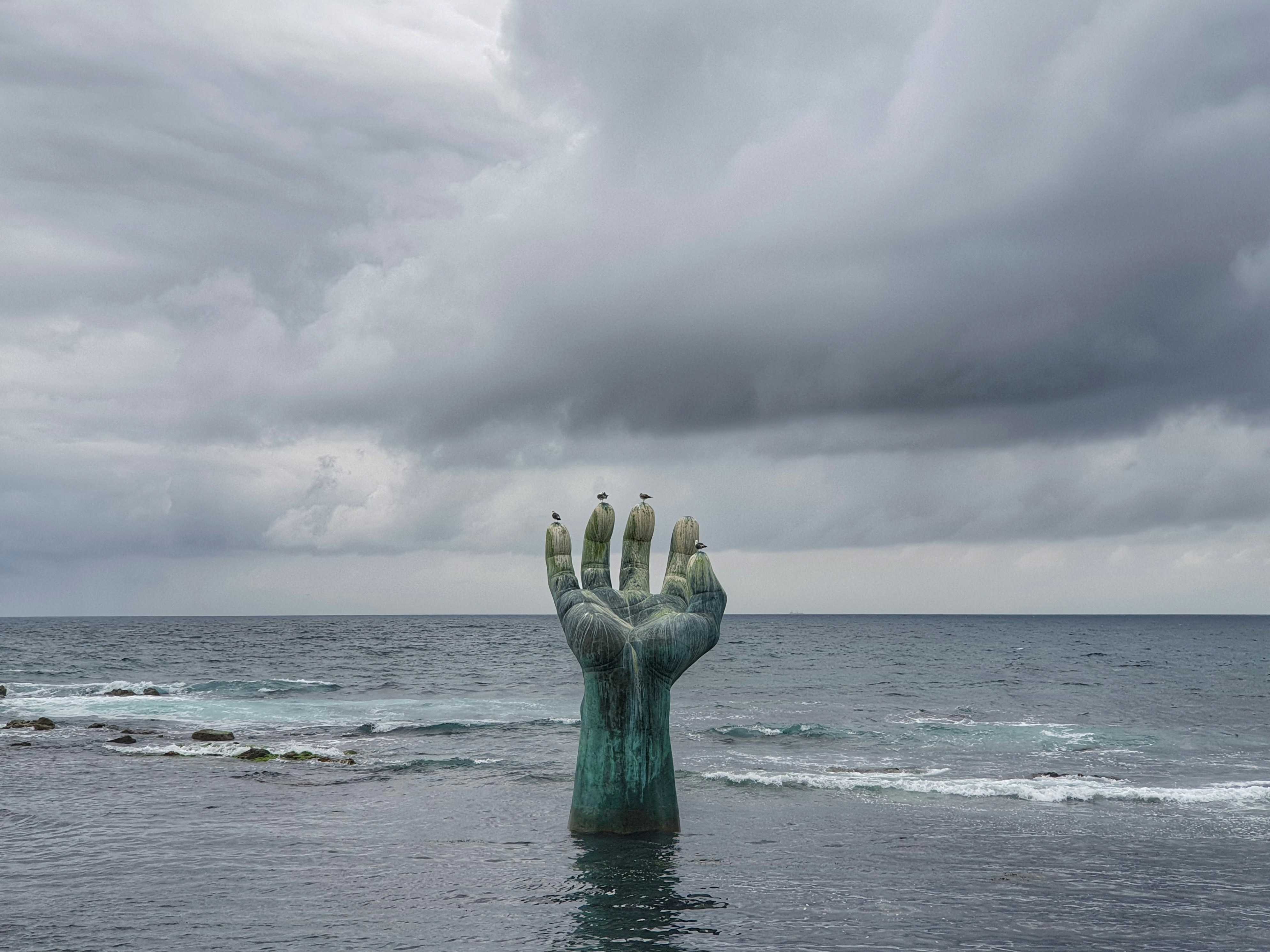 A large, weathered hand sculpture rises from the ocean, symbolizing resilience against a backdrop of dramatic clouds and waves.
